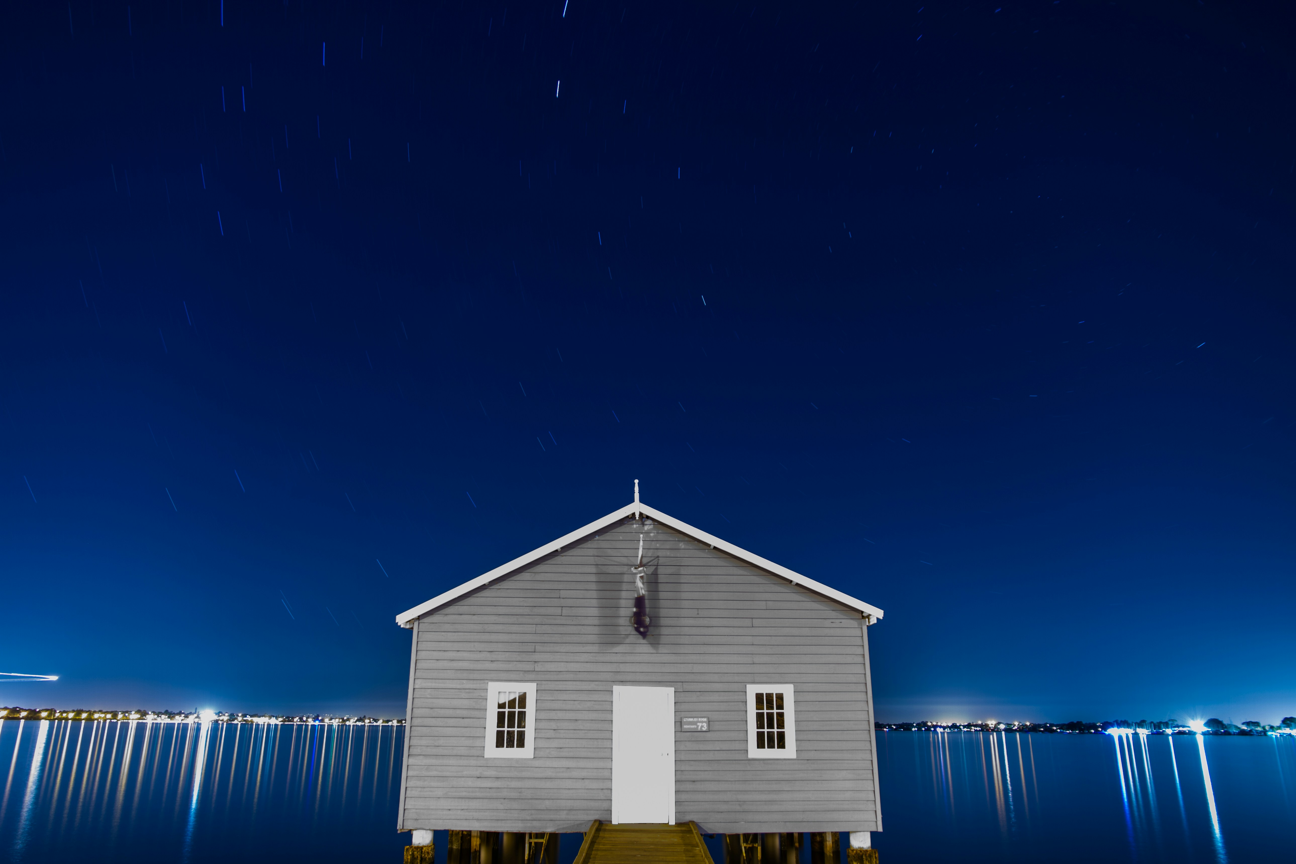 Wooden boathouse on a pier under a clear, starry night sky with reflections on calm water.