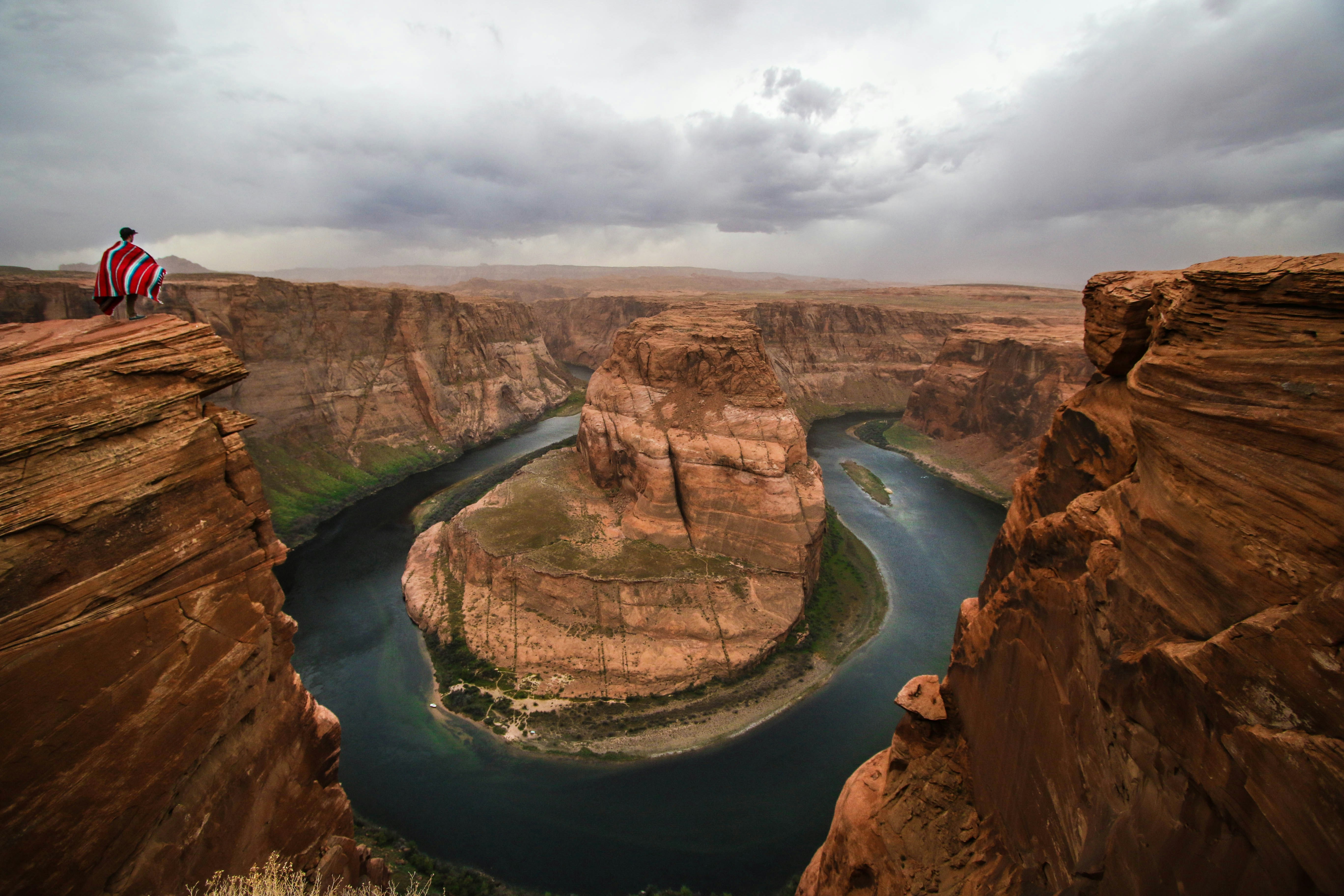 Person wrapped in a colorful blanket sits on a cliff edge overlooking the dramatic curve of Horseshoe Bend under a cloudy sky.