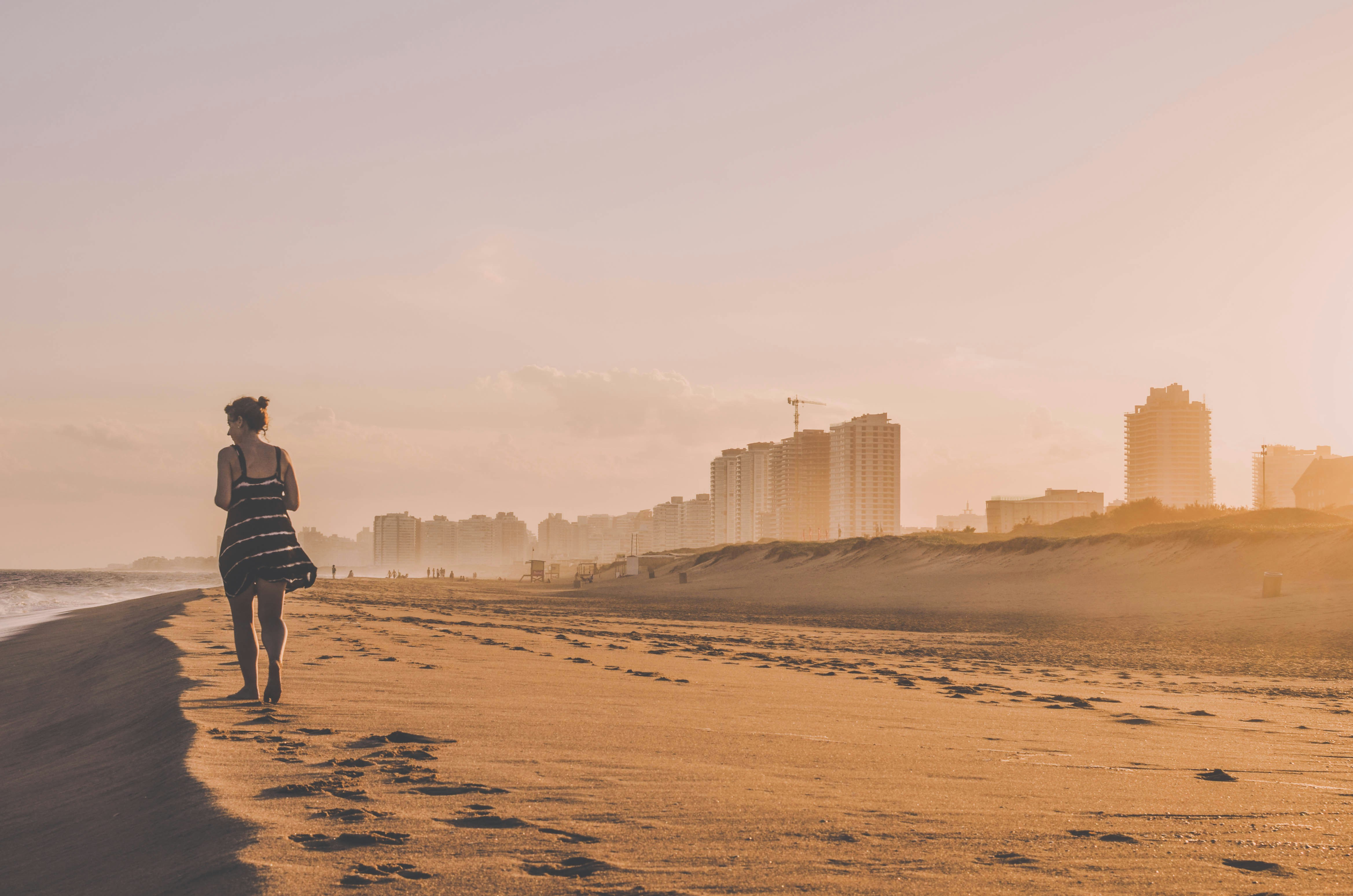 A woman strolls along a sandy beach at sunset, with city skyscrapers softly silhouetted in the background. The scene evokes a sense of tranquility and urban contrast.