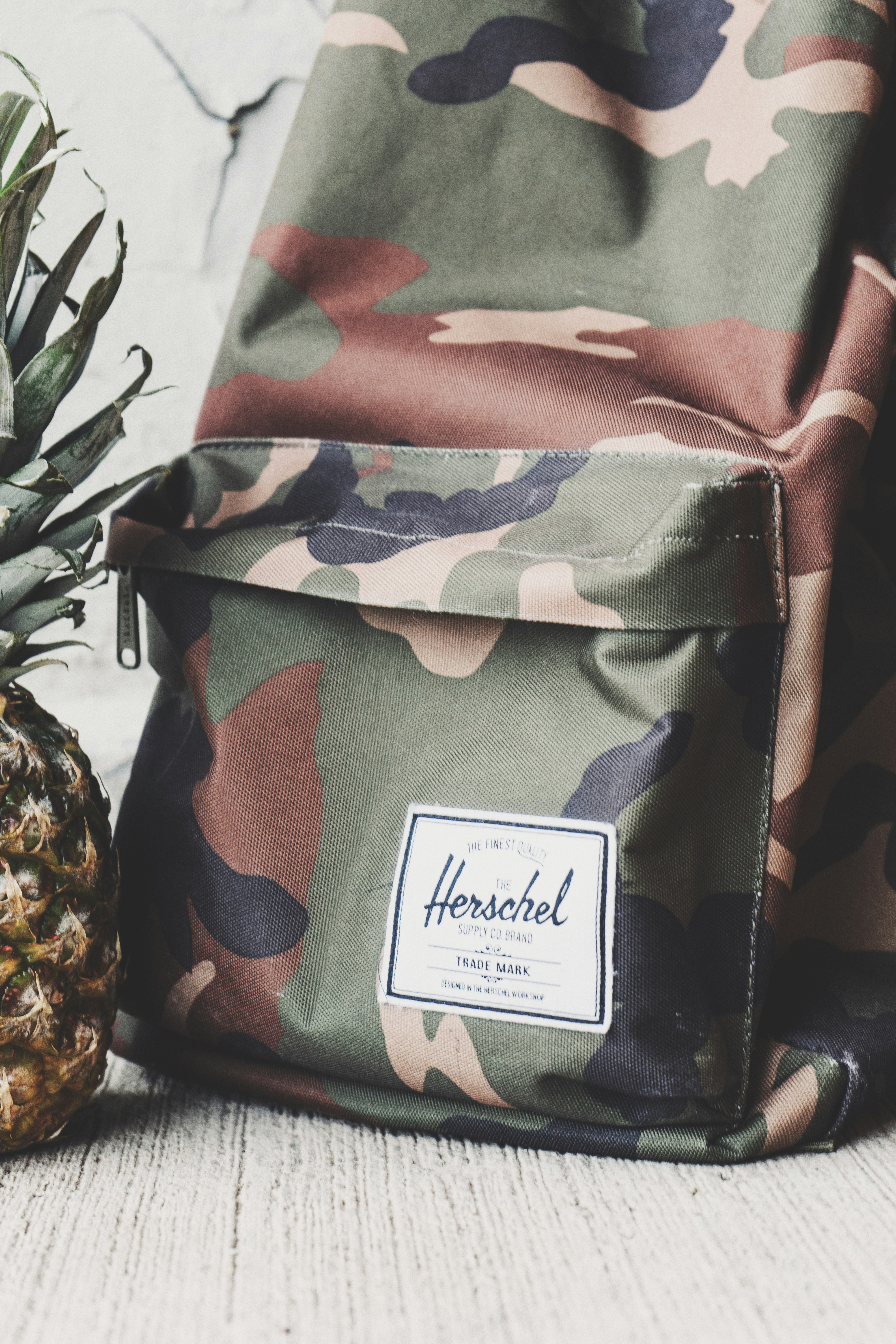 Camo backpack with Herschel logo beside a pineapple on a textured surface.