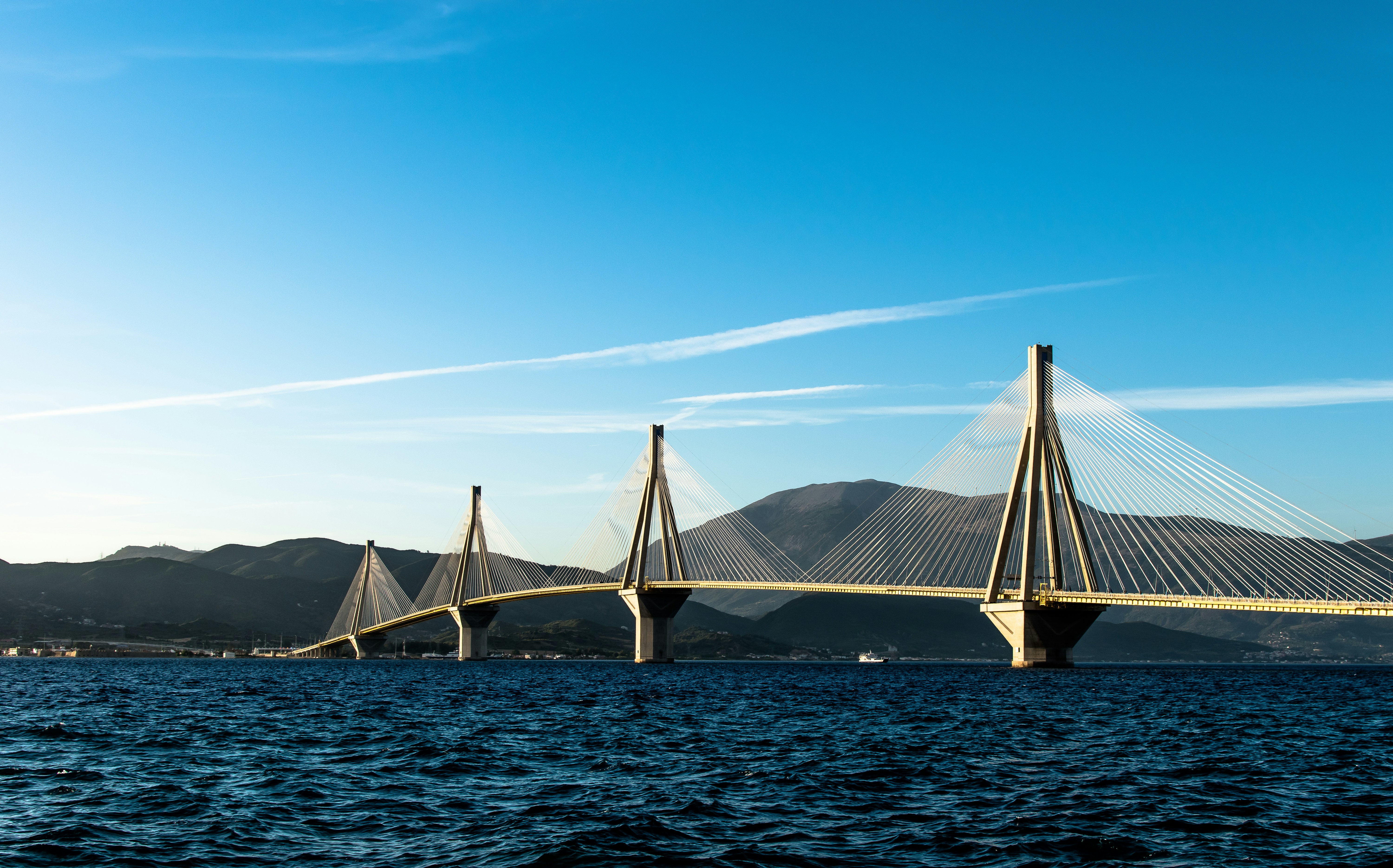 Elegant cable-stayed bridge spanning a tranquil body of water, framed by distant mountains under a clear blue sky.