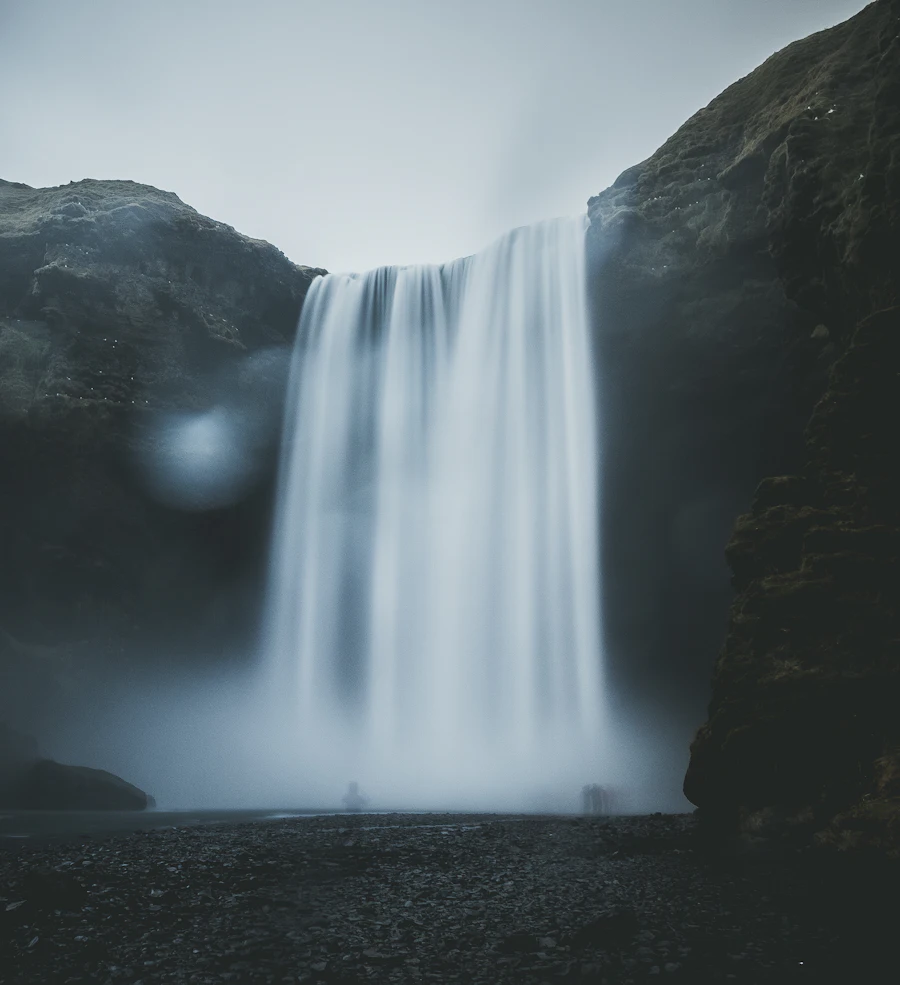 A broad waterfall pours between mossy dark cliffs into a misty basin, with silky blurred water and a moody gray sky overhead.