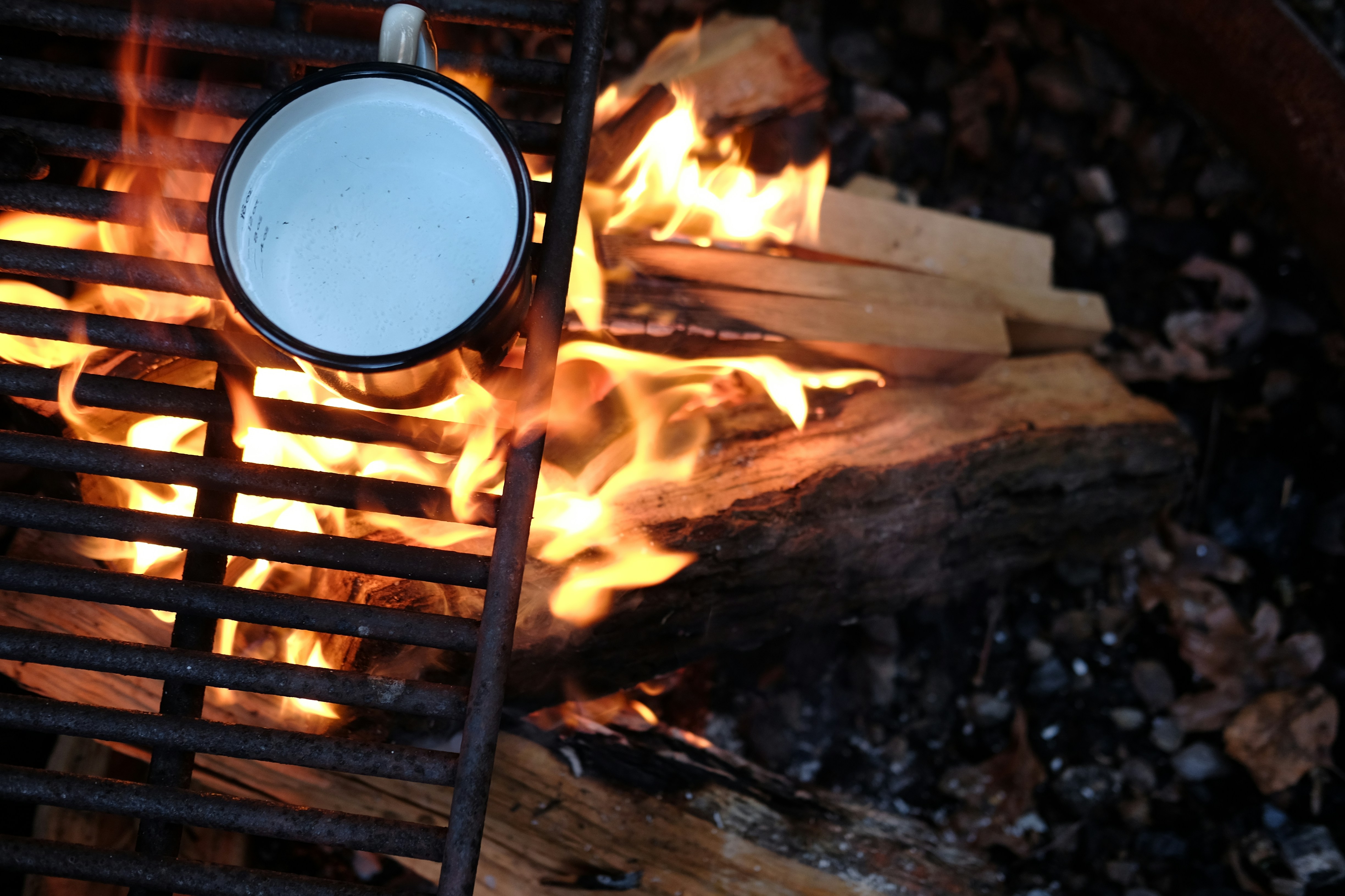 A metal mug filled with water sits on a grill above a crackling campfire, surrounded by glowing embers and charred wood.
