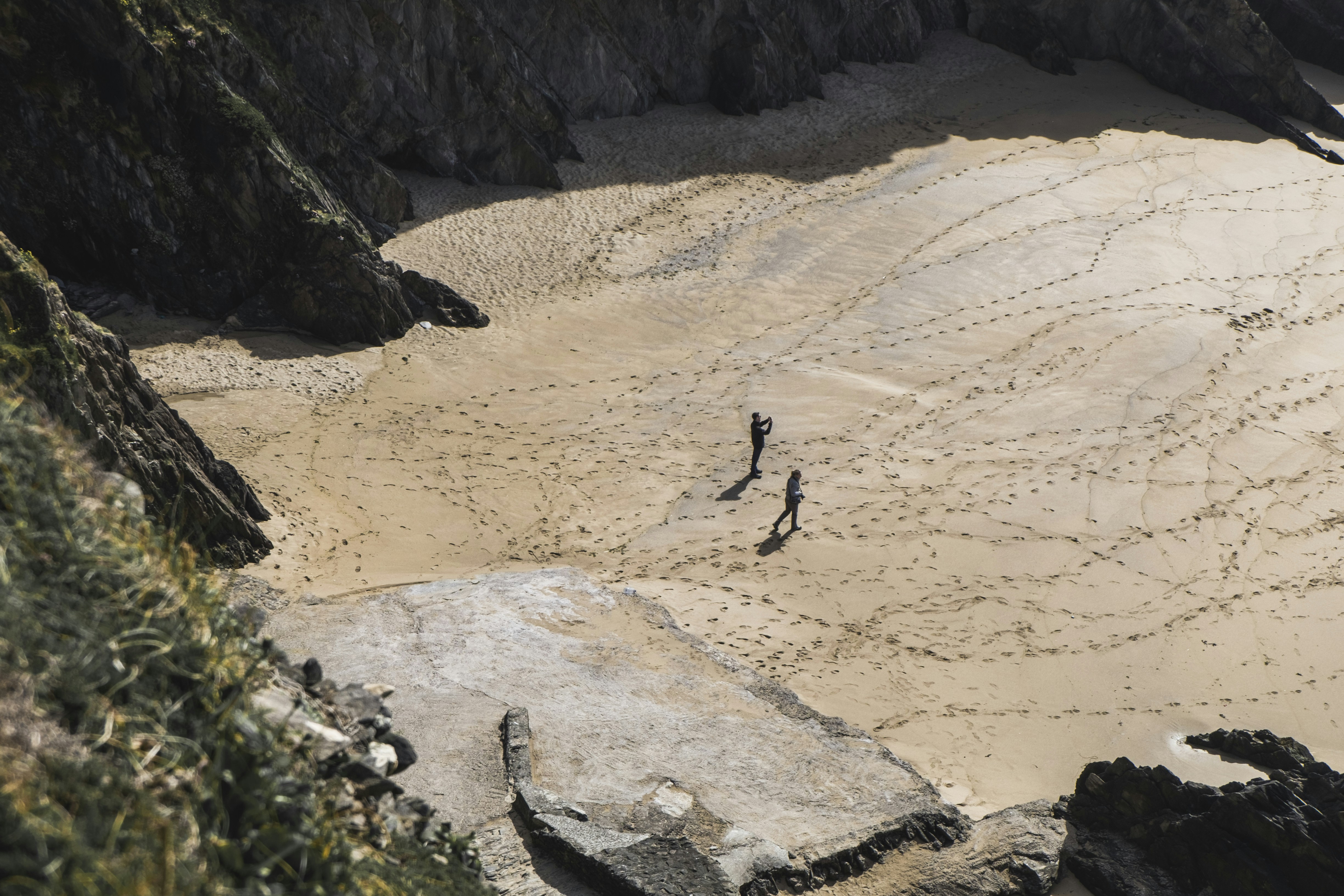 Two figures walk across a secluded, footprint-marked beach surrounded by rugged cliffs.