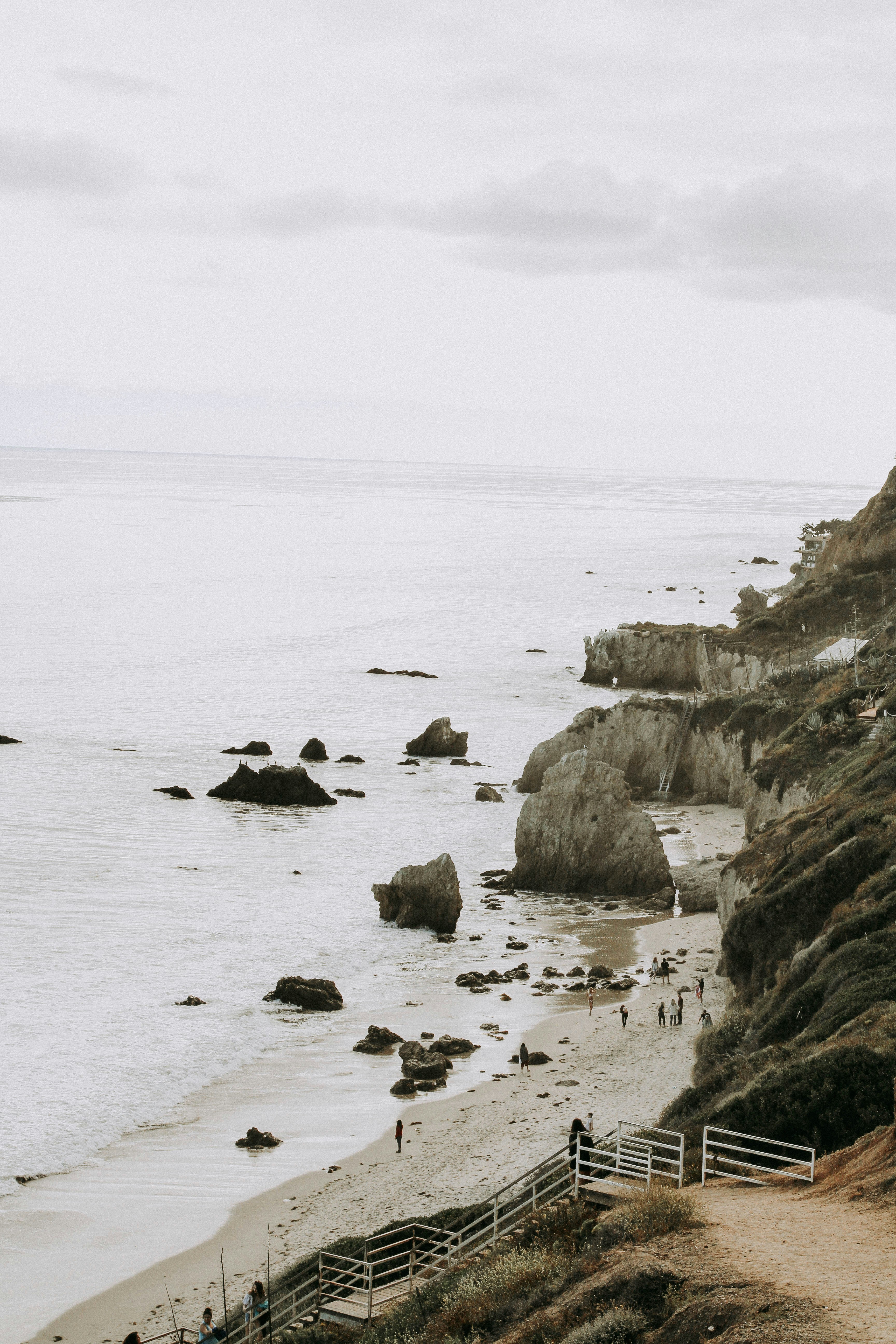 People walking along a sandy beach with rocky cliffs under a cloudy sky, capturing the tranquility of a coastal landscape.