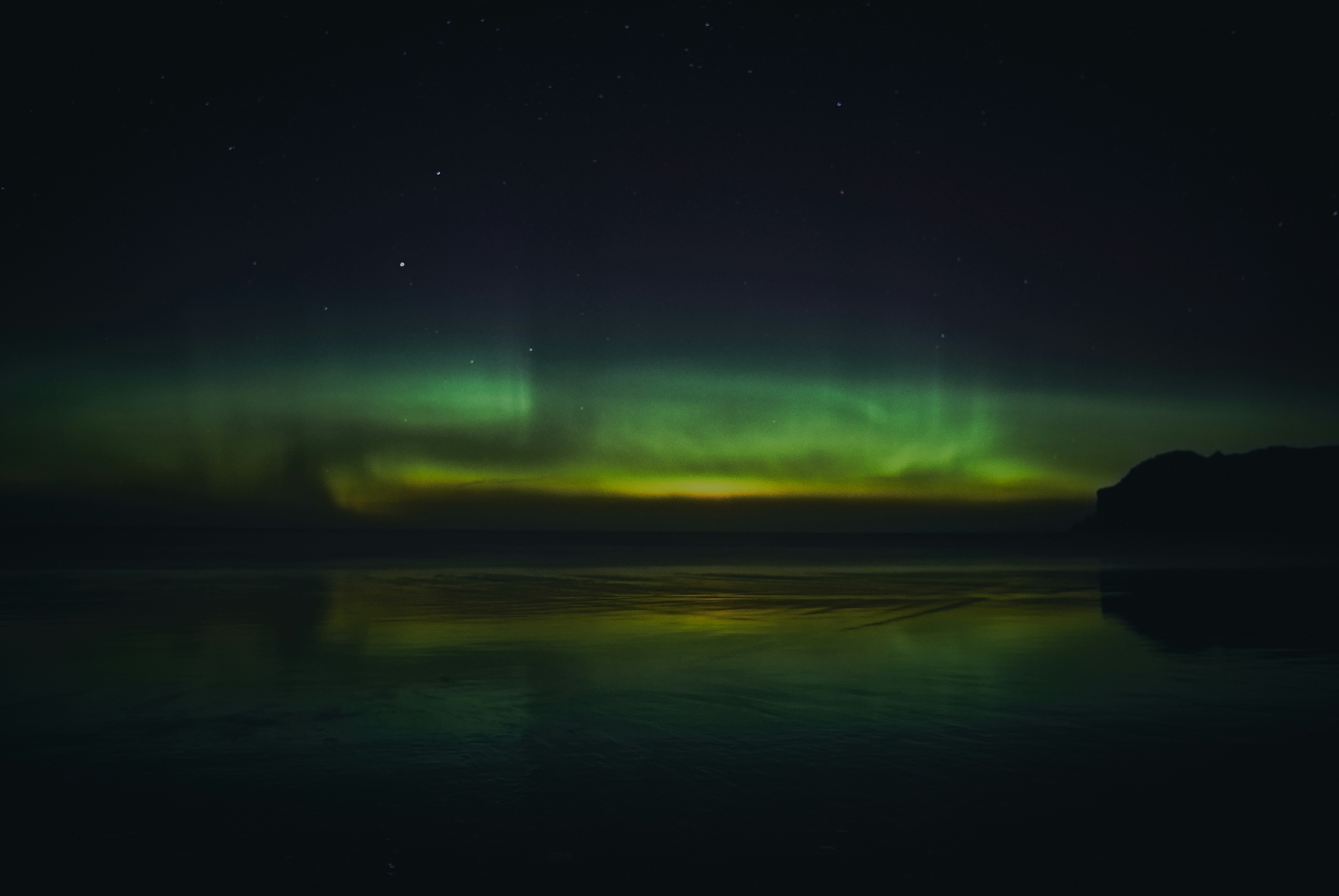 Green aurora lights shimmer over a calm, dark sea with distant silhouetted land.