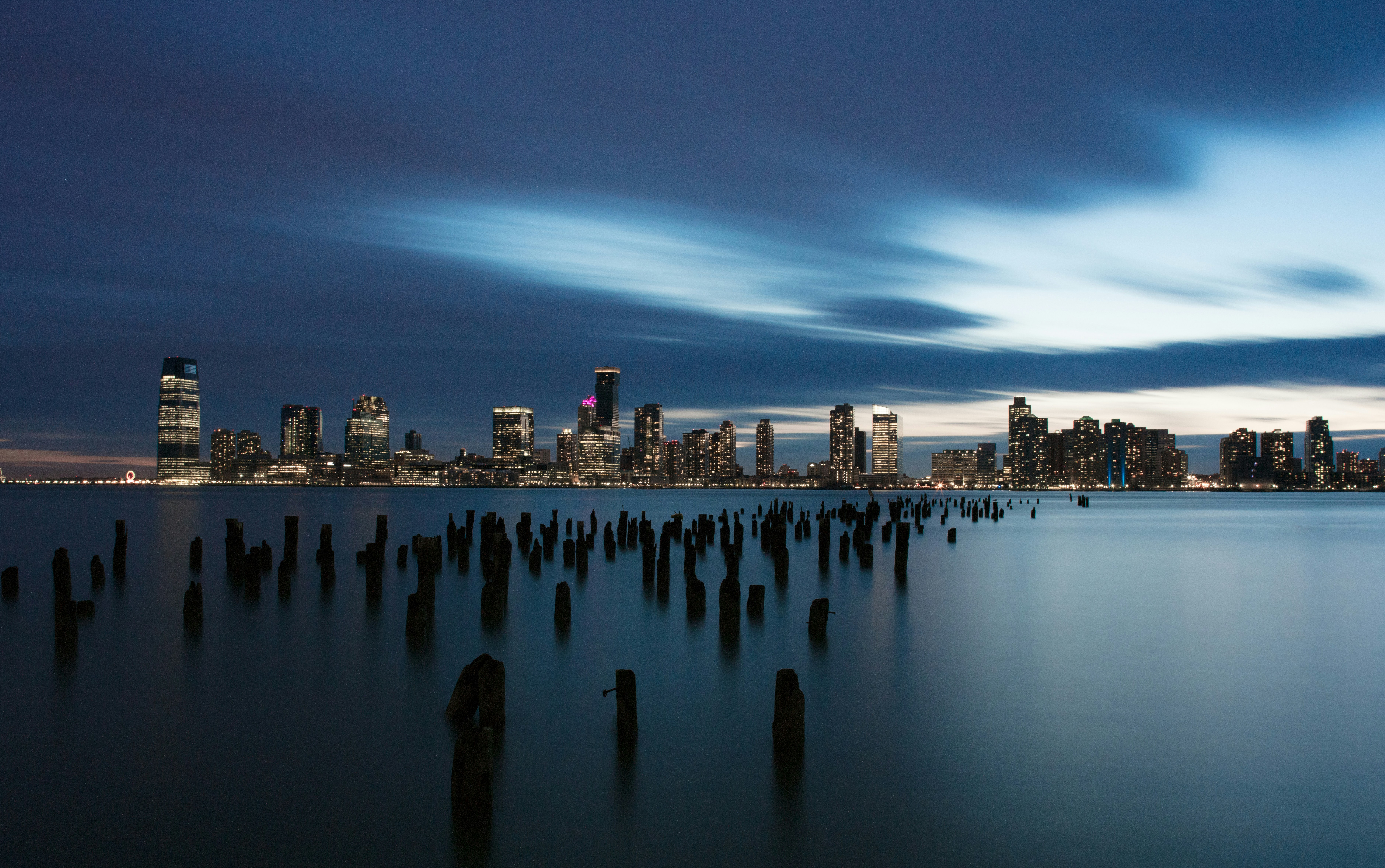 City skyline at dusk with silhouetted wooden posts in the foreground and a blue sky overhead.