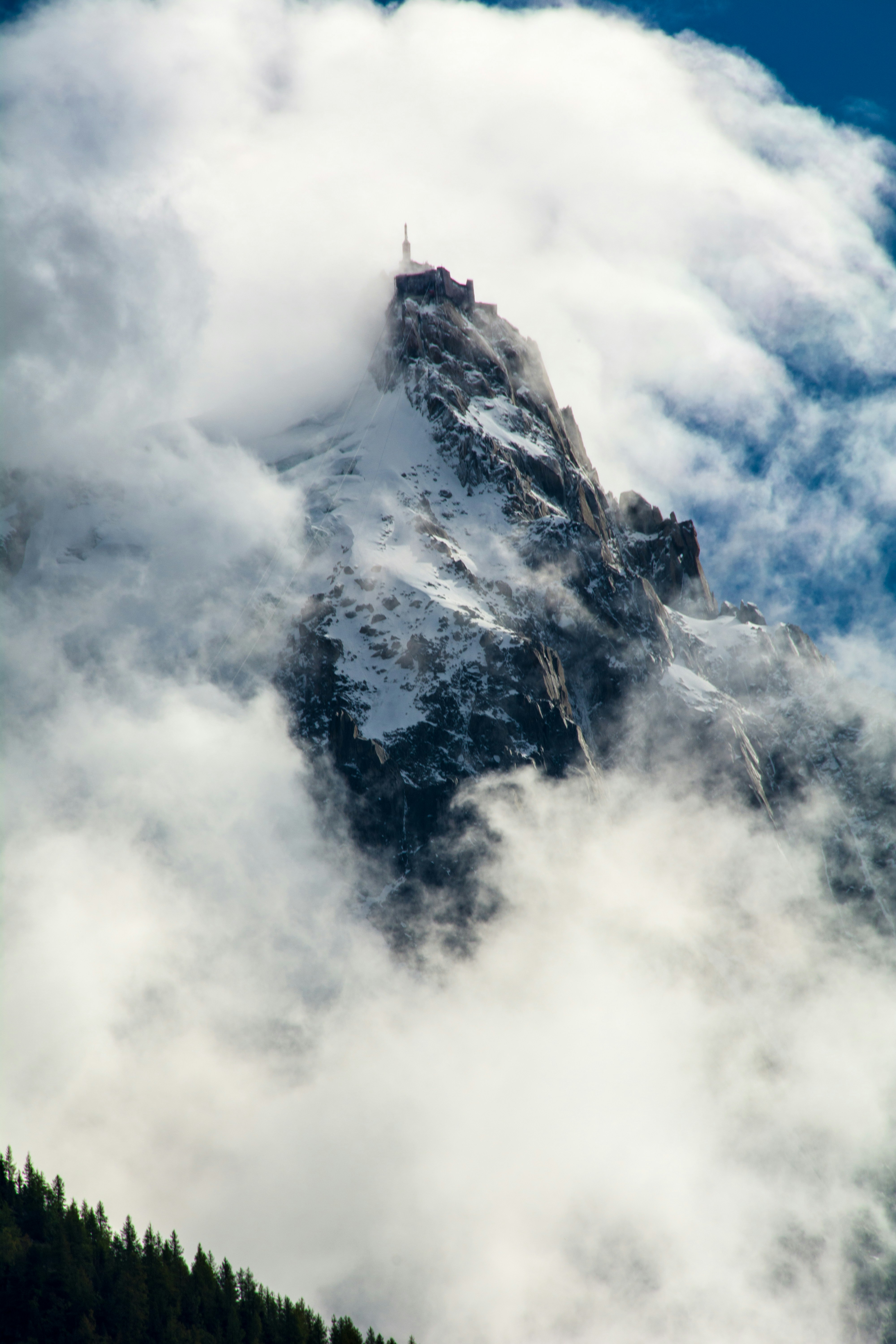 Snow-capped mountain peak partially shrouded in clouds, revealing a distant structure at the summit. The scene captures the majesty of nature and human endeavor.