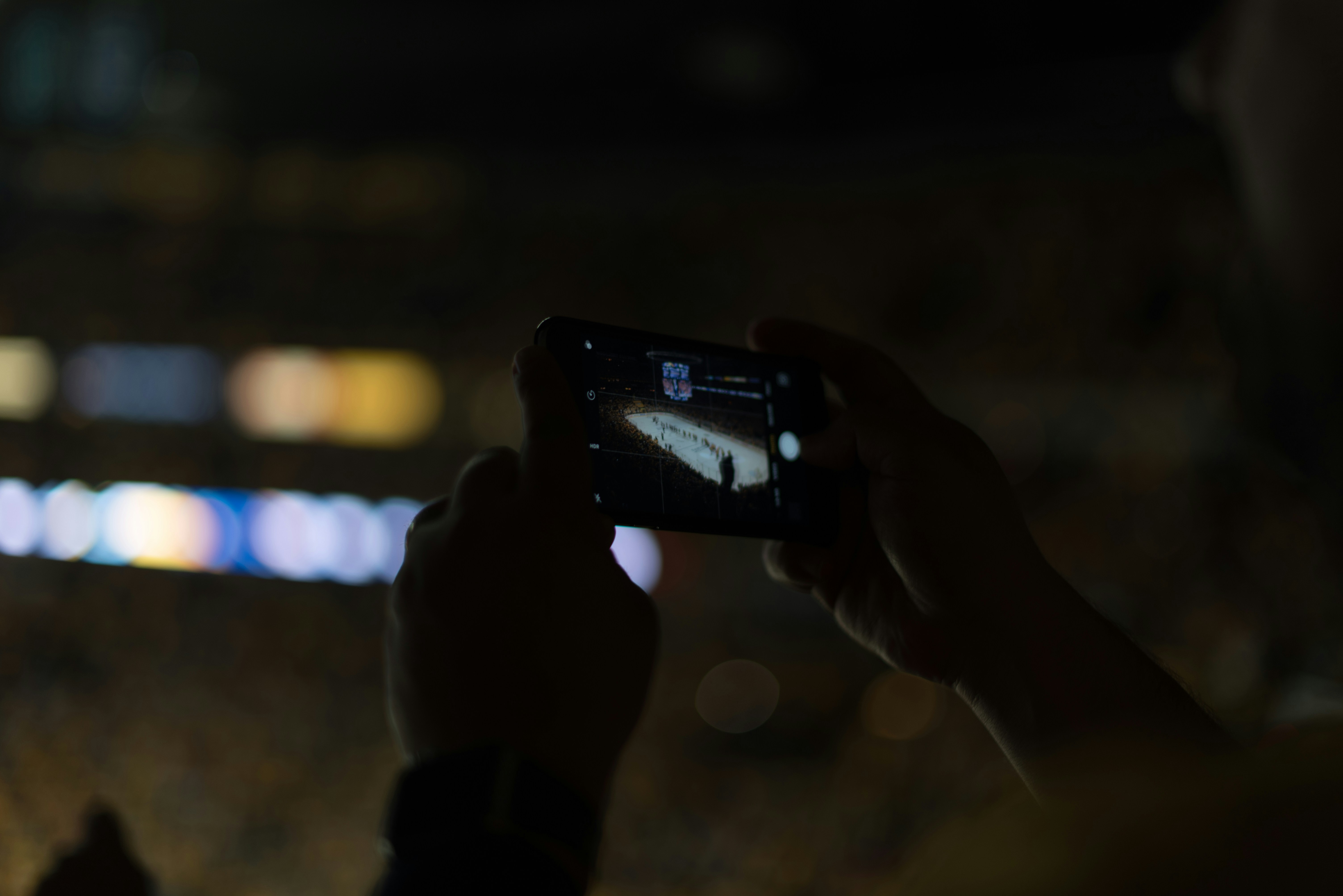 Silhouetted smartphone capturing a distant sports field under dim stadium lights.