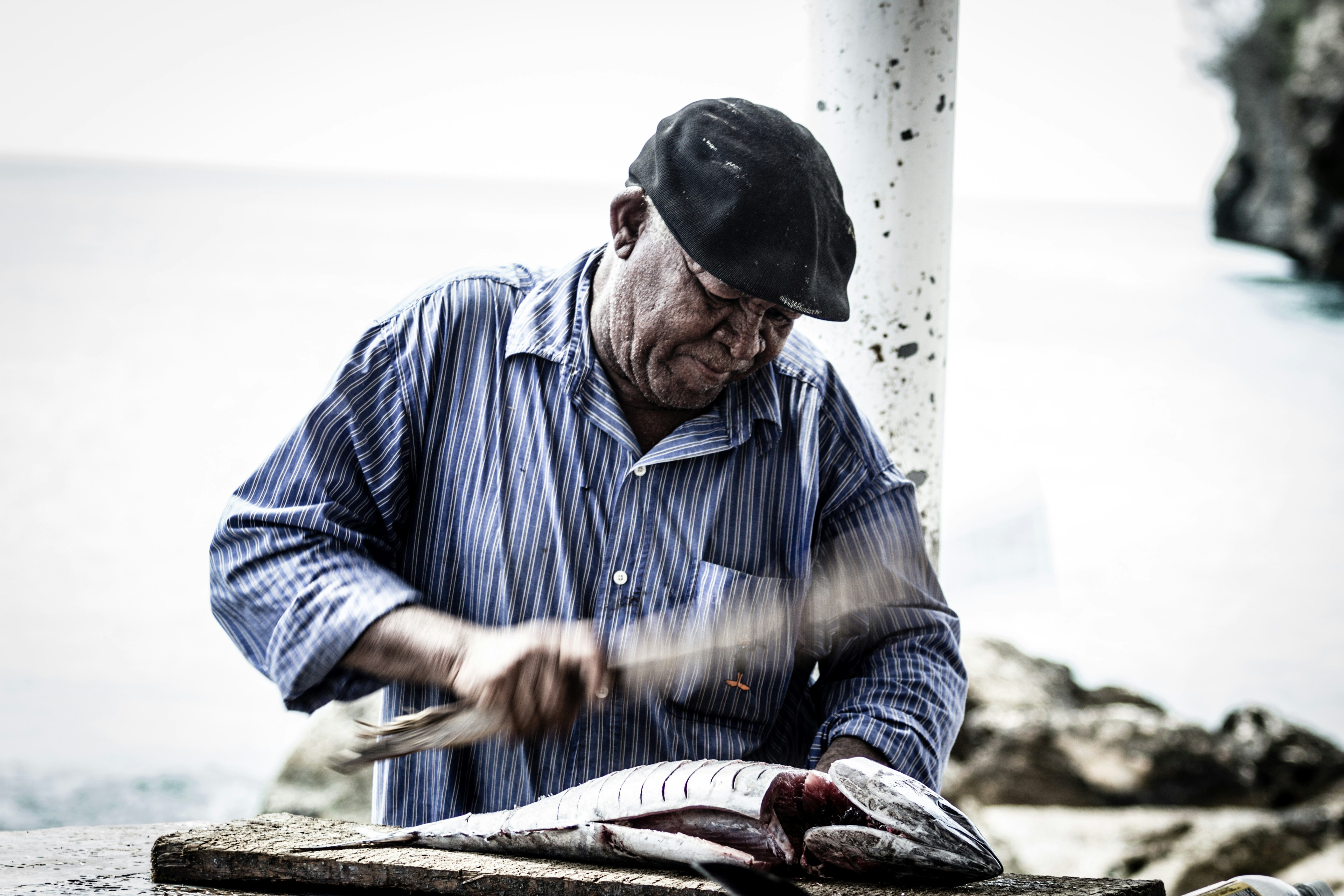 An elderly man skillfully filleting a fish at a coastal setting, showcasing traditional culinary techniques. The serene ocean backdrop enhances the scene's authenticity.