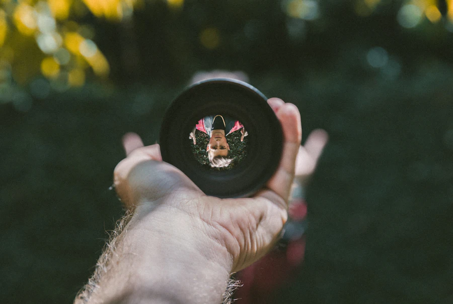 A hand holds a camera lens toward the viewer, with a small upside-down figure reflected inside the glass. Soft green and gold bokeh fills the background.