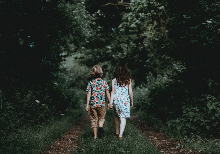 Two children holding hands while walking down a lush, green path surrounded by dense foliage.