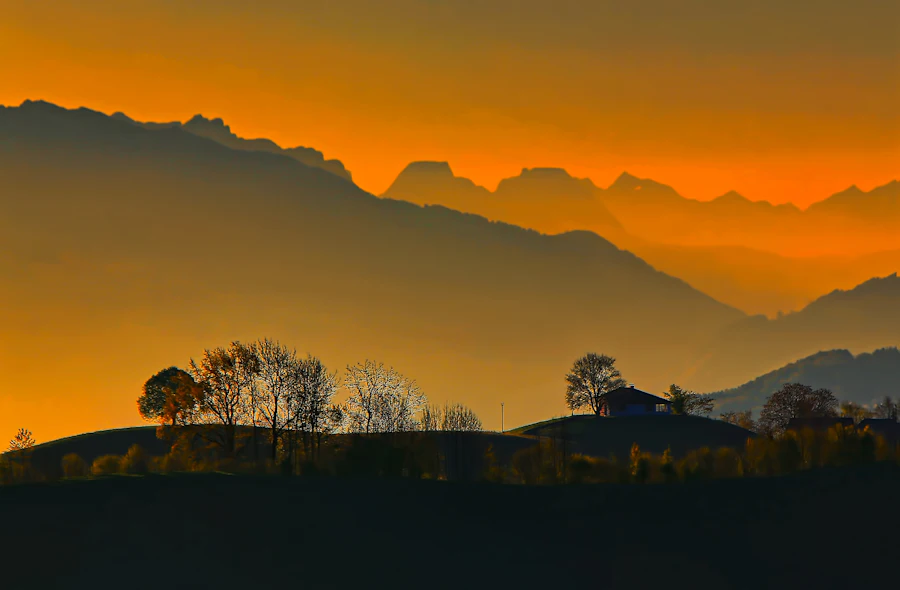 Layered mountain ridges glow in orange dawn light above a dark foreground hill with silhouetted trees and a small house. The sky fades from deep amber to soft haze.