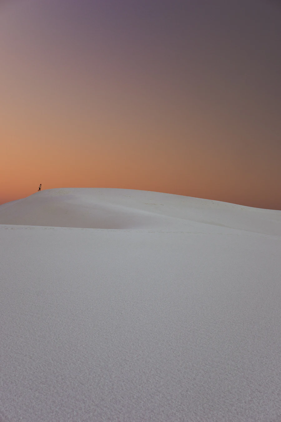 A lone figure stands atop a smooth, white dune under a gradient sky transitioning from soft orange to deep purple.
