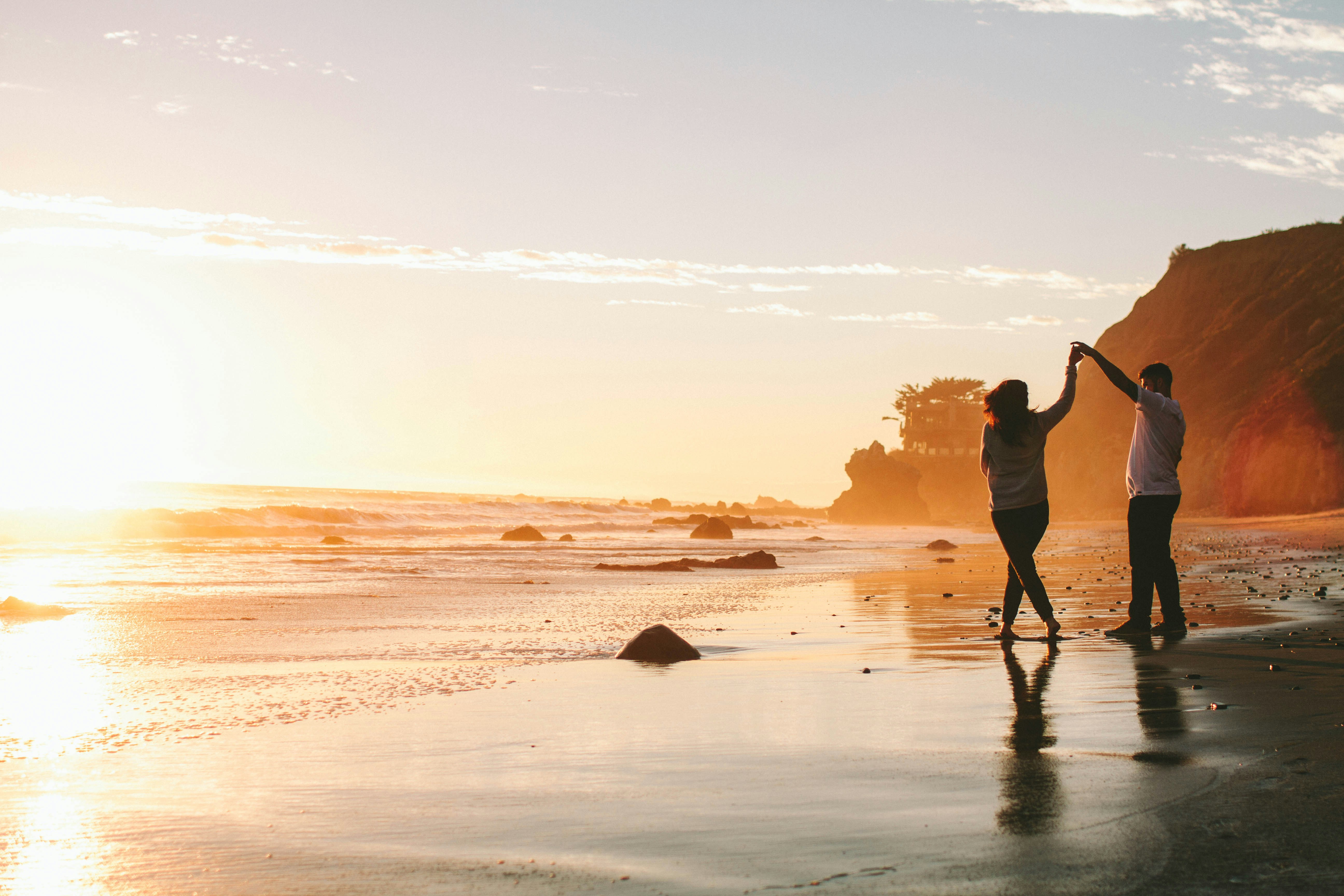 Couple dancing on a sandy beach during a golden sunset, with waves gently lapping the shore.