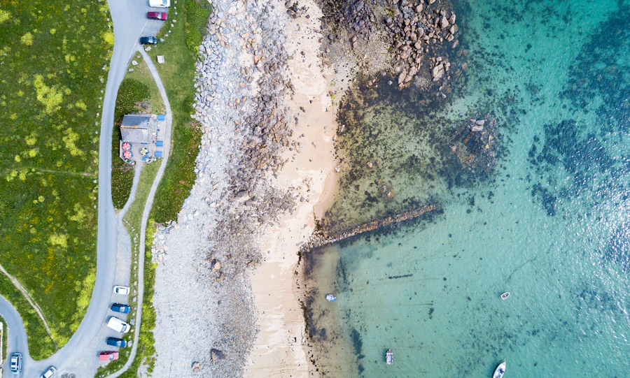Top-down view of a small beach cove with pale sand, dark rocks, and clear turquoise water beside a winding coastal road and parked cars.