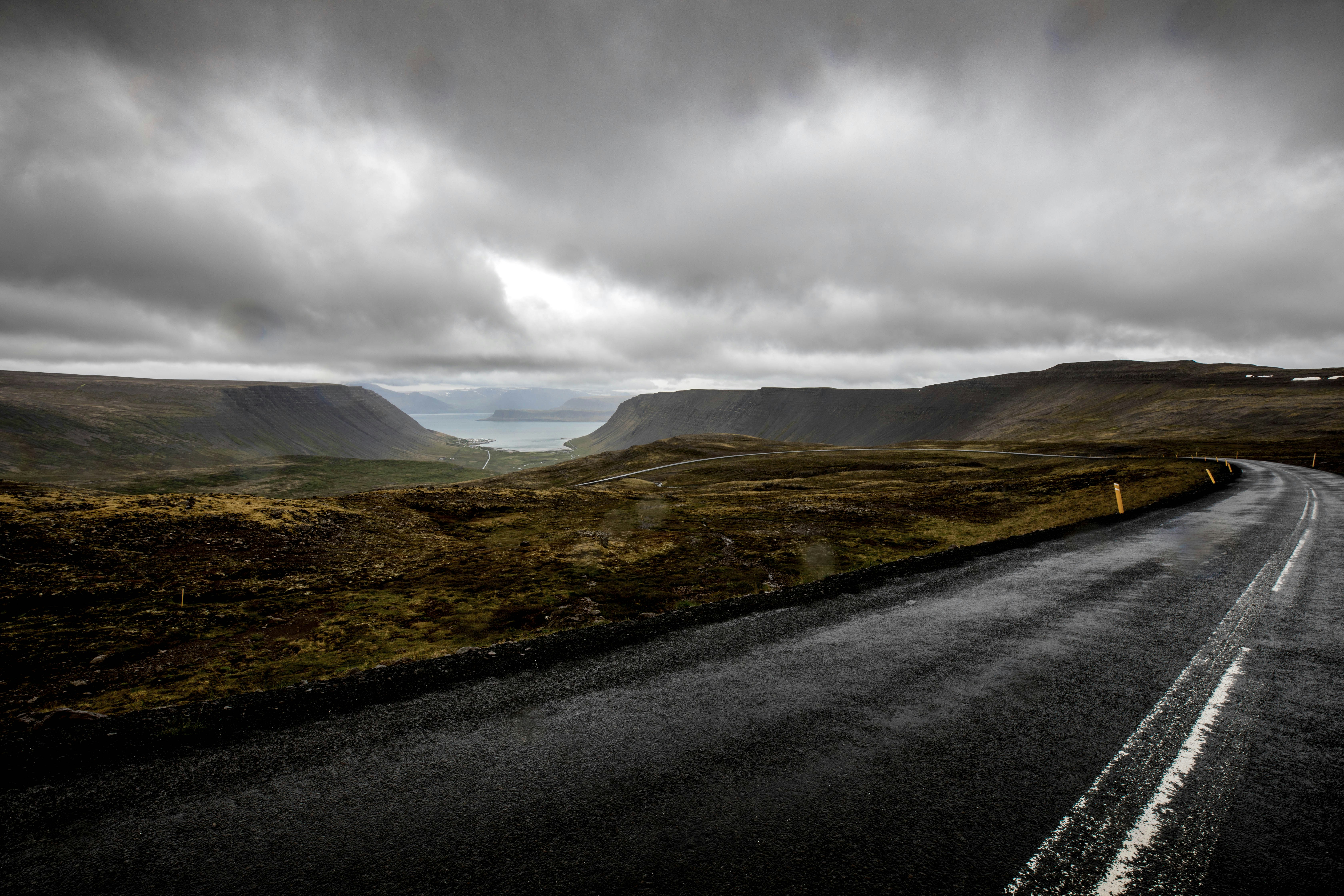 Wet asphalt road winding through a mountainous landscape under a cloudy sky.