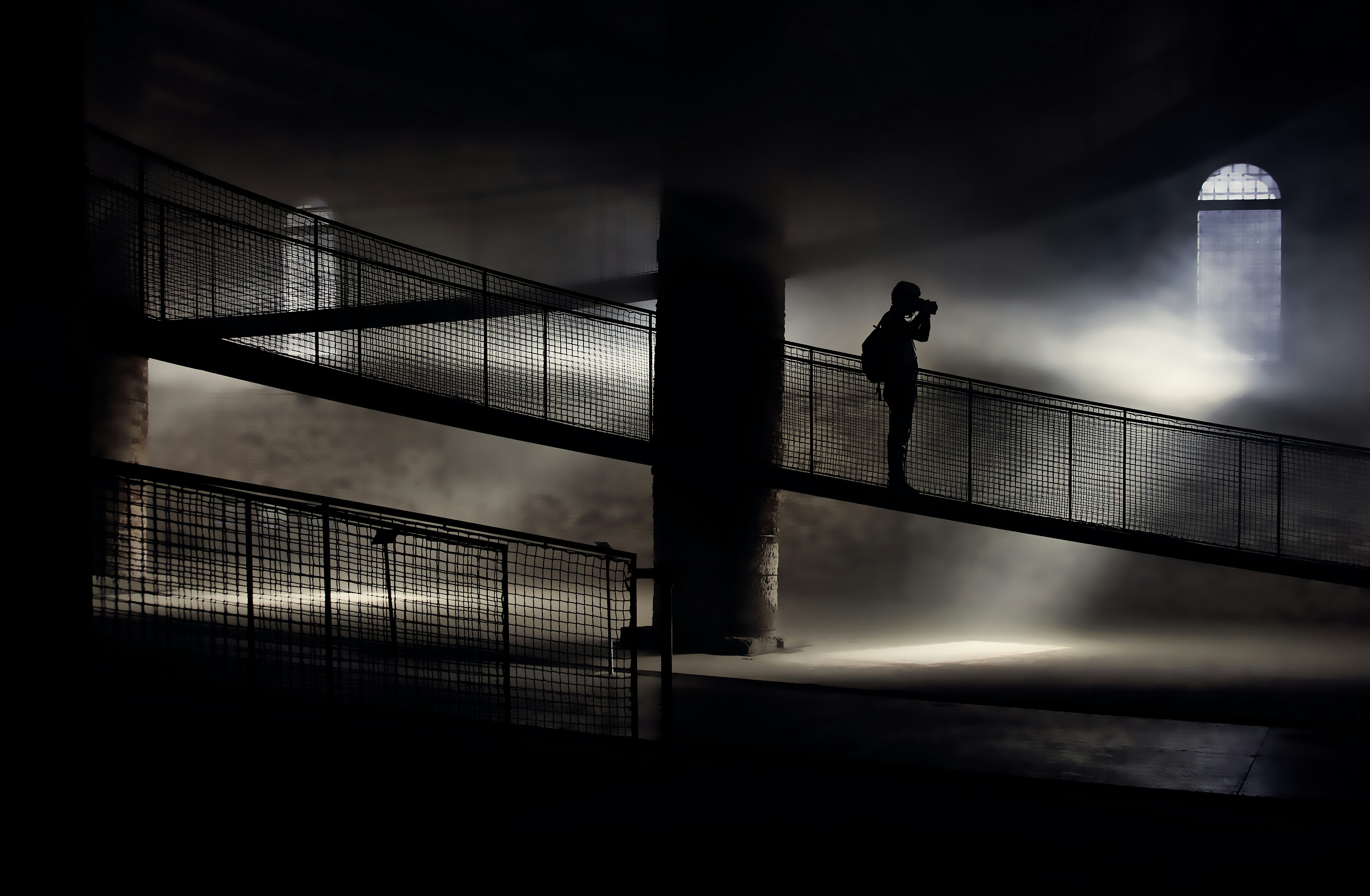 A lone silhouette stands on a fog-blanketed catwalk, backlit by a distant arched window.