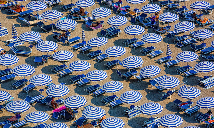 High-angle view of a crowded beach filled with blue-and-white striped umbrellas and matching sun loungers on the sand. A few people are scattered among the neatly arranged rows.