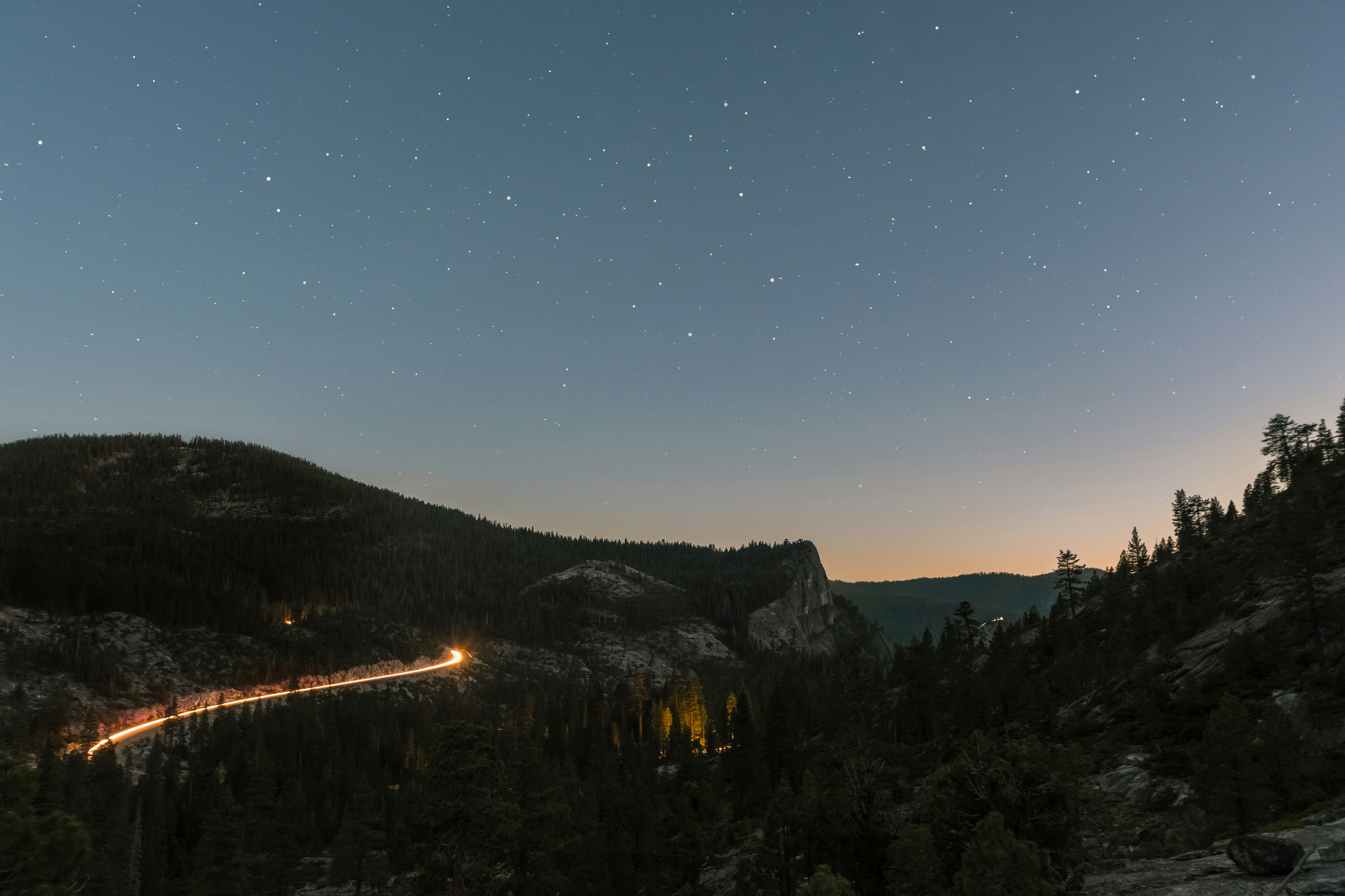 Starry night sky over a mountain landscape with a winding road illuminated by car lights.