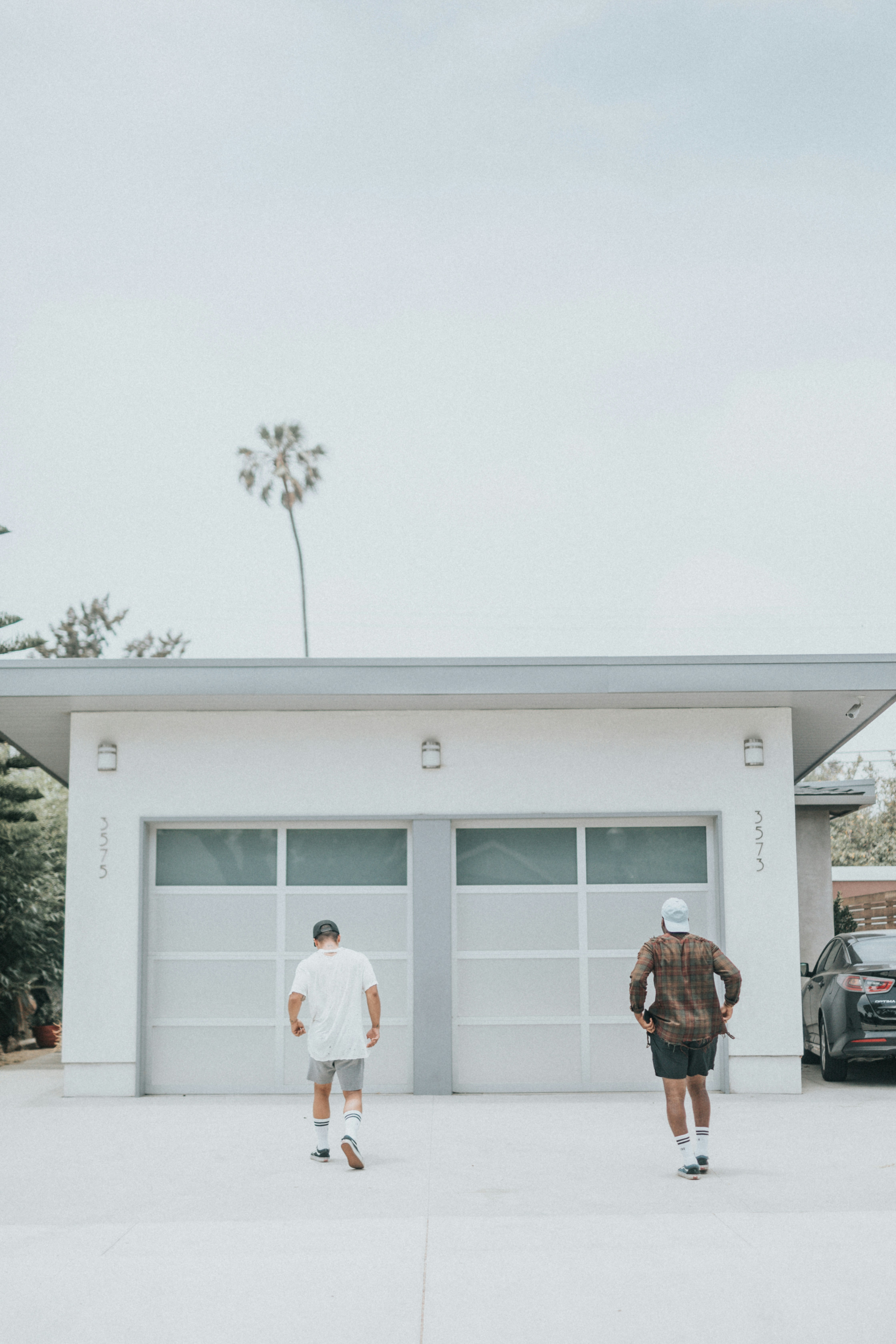 Two individuals walking away from a modern house with a minimalist design, framed by palm trees and a sleek car.