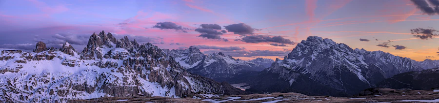 Snowy mountain range under a pink and purple sunset sky, with rugged peaks stretching across the horizon and dark valleys below.
