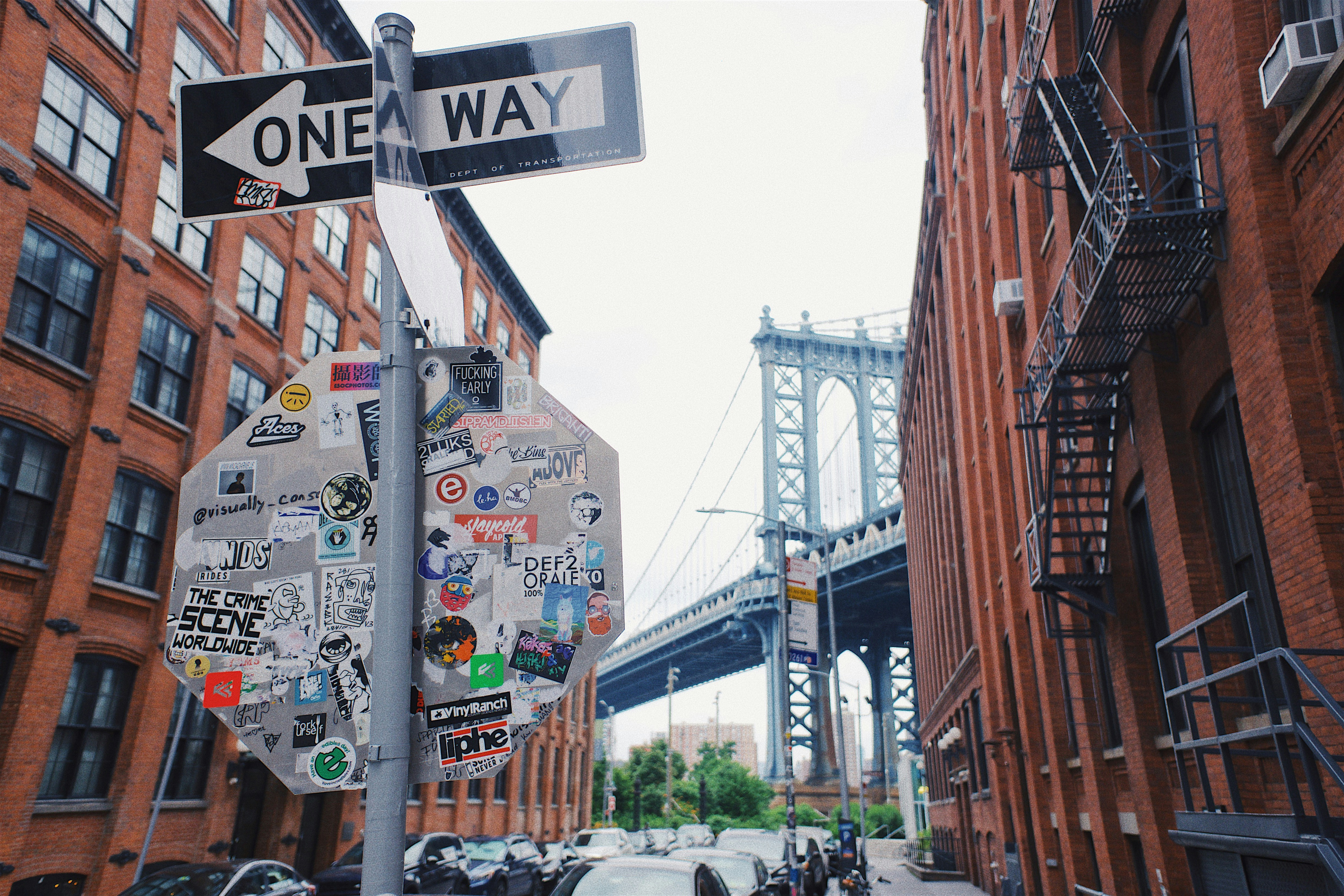 A street sign adorned with stickers stands in the foreground, framed by the iconic Manhattan Bridge and red brick buildings in Brooklyn.
