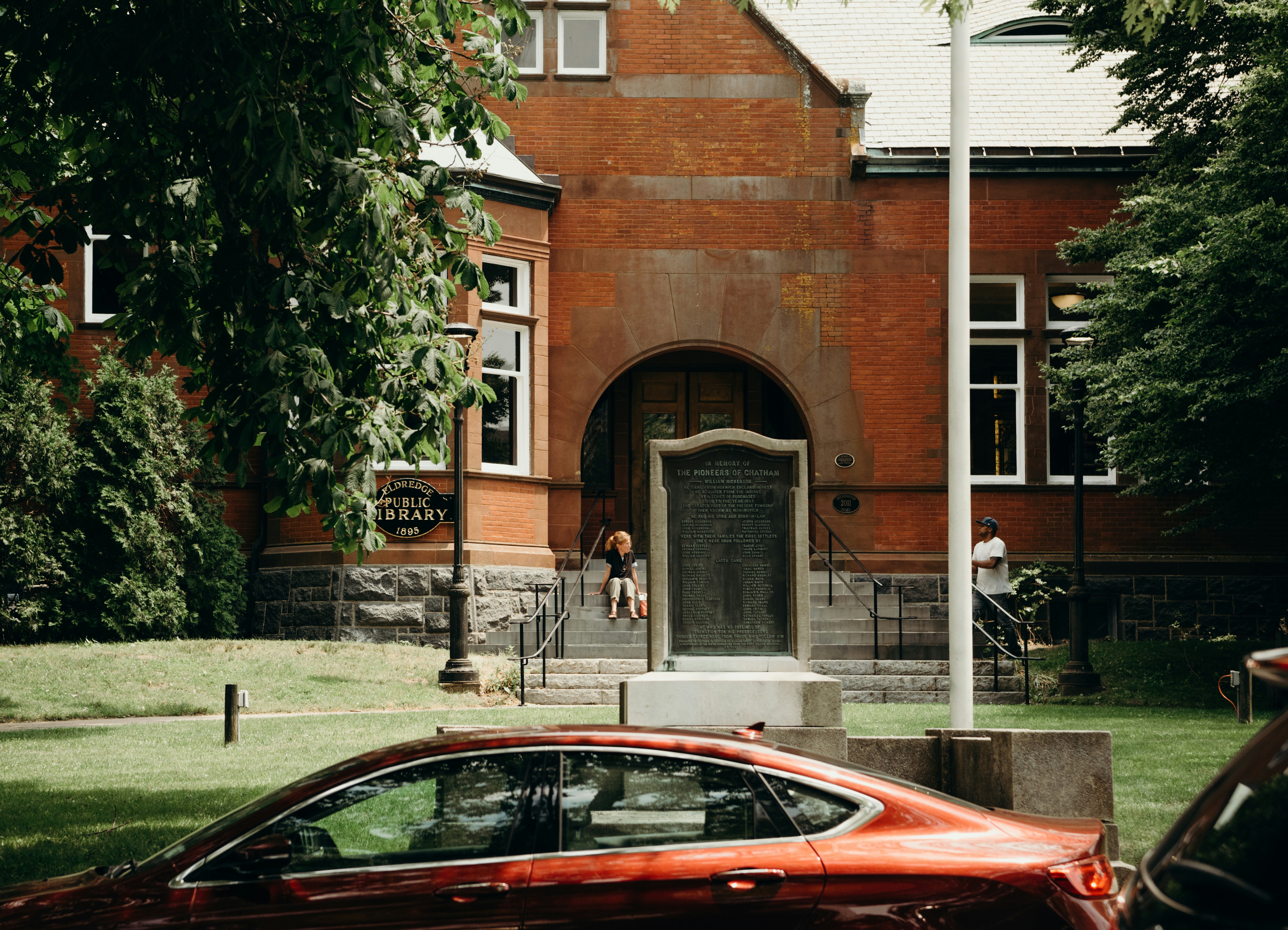 Historic library building with a commemorative monument in front, surrounded by lush greenery and a parked red car.