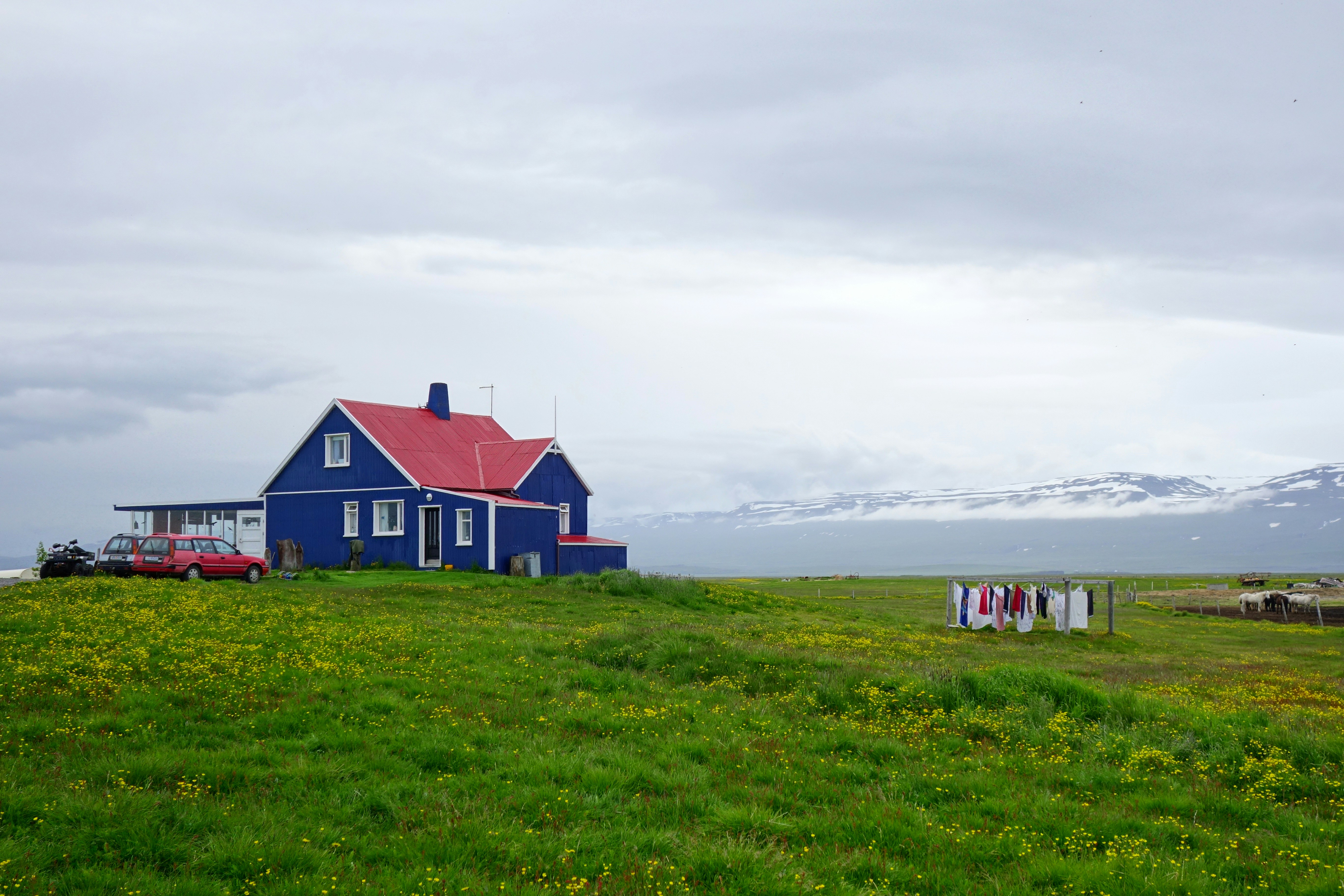 Colorful blue house with a red roof set against a backdrop of mountains and green fields, complemented by clothes hanging on a line. 