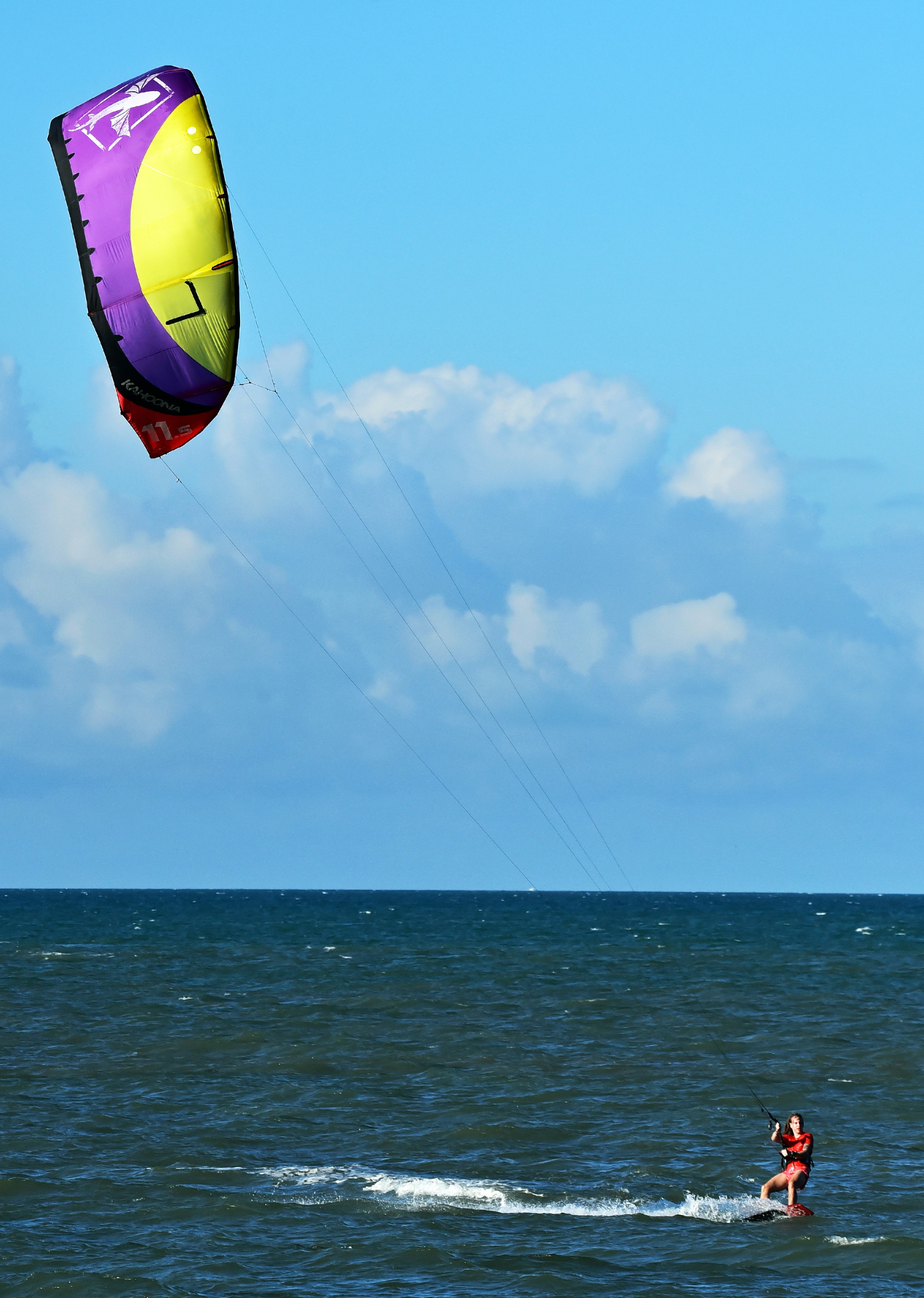 A kite surfer skillfully maneuvers across the ocean with a vibrant kite soaring above. The scene captures the thrill of water sports against a backdrop of fluffy clouds.