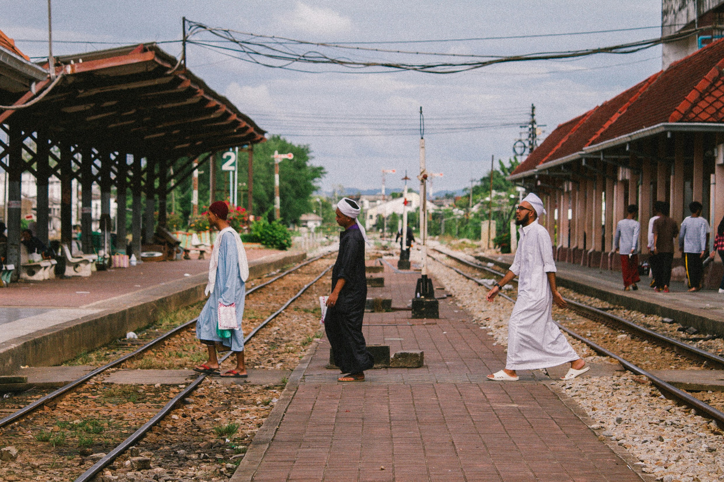 Three men in traditional attire cross the tracks at Yala railway station under a cloudy sky.