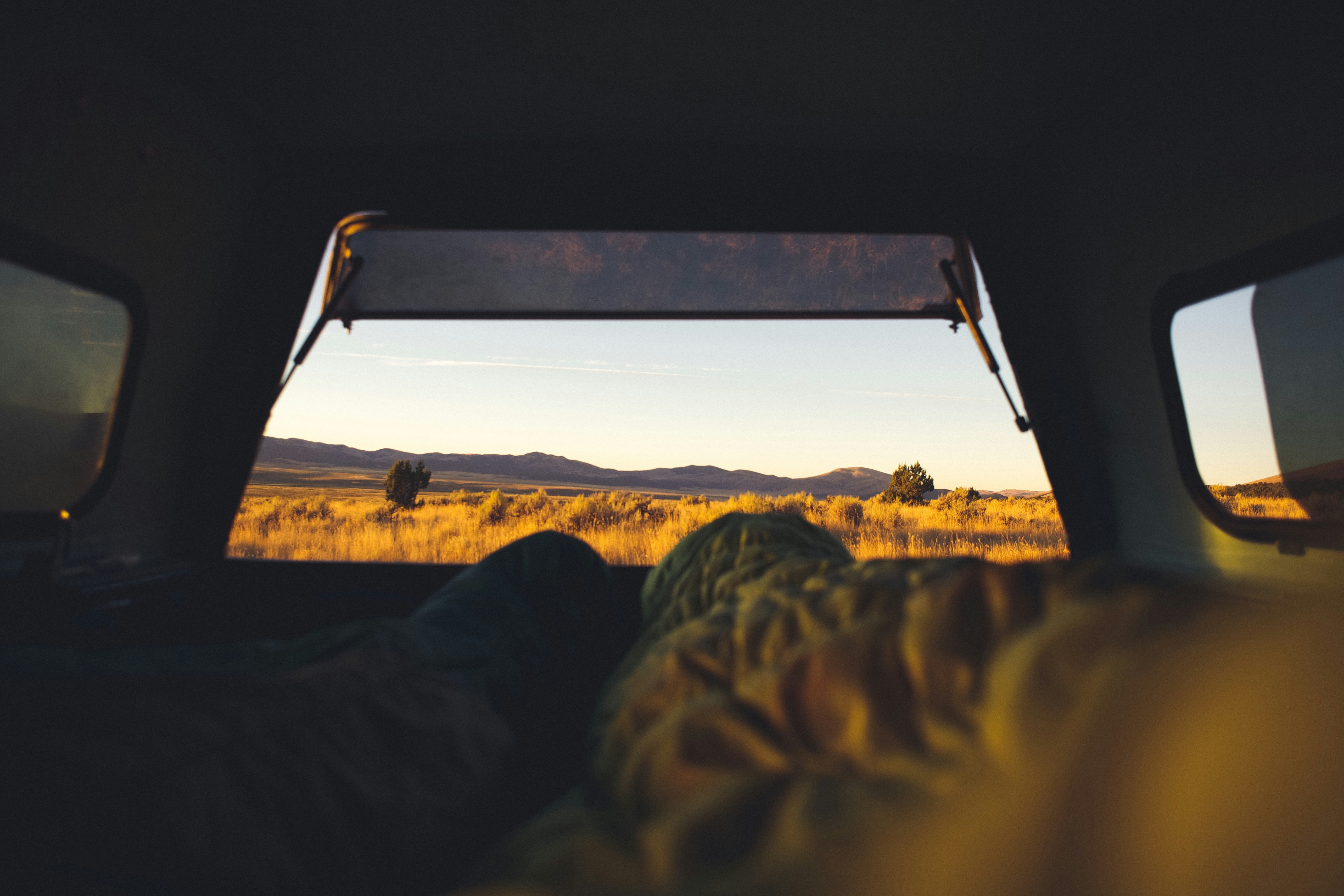 View of golden grasslands and distant mountains framed by the open rear window of a camper, suggesting a tranquil morning escape.