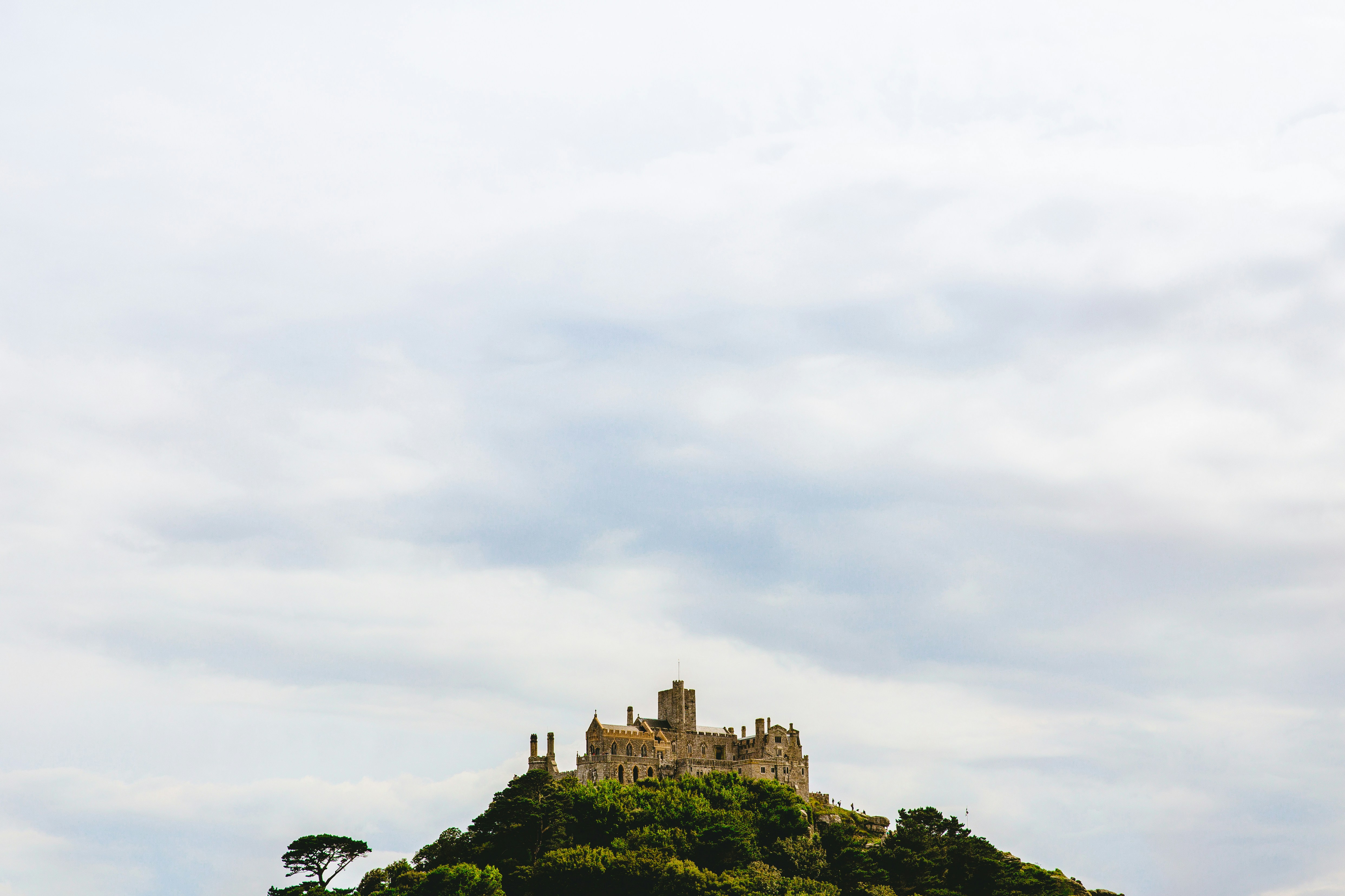 Historic castle perched atop a lush green hill under a wide expanse of overcast sky.