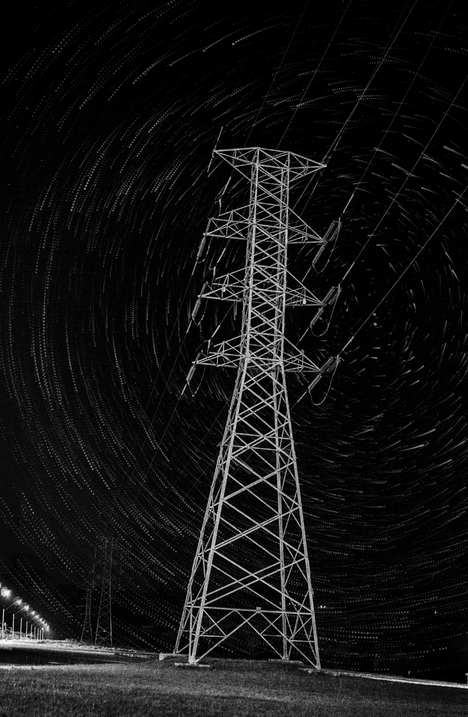 A towering power line stands against a swirling night sky, captured in a long exposure that reveals dynamic star trails. The contrast of the structure against the celestial background creates a striking visual narrative.