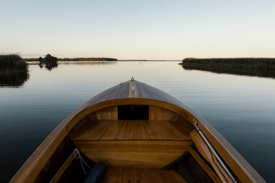 Wooden boat bow pointing across a calm lake at dusk, with reed-lined shores on both sides and a pale sky reflected on the surface.