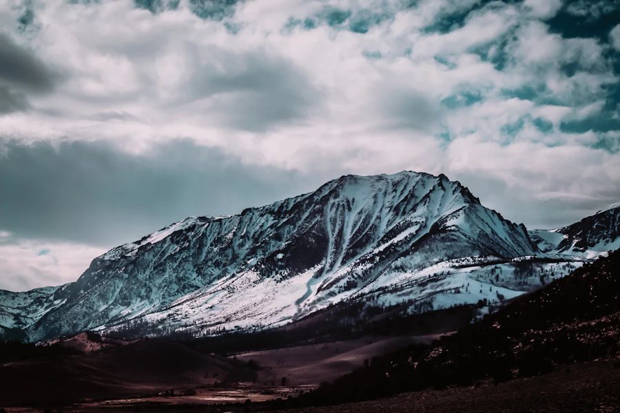 Snow-covered mountain ridge rising above a dark valley beneath a sky filled with layered clouds. The peak and snow gullies dominate the scene.