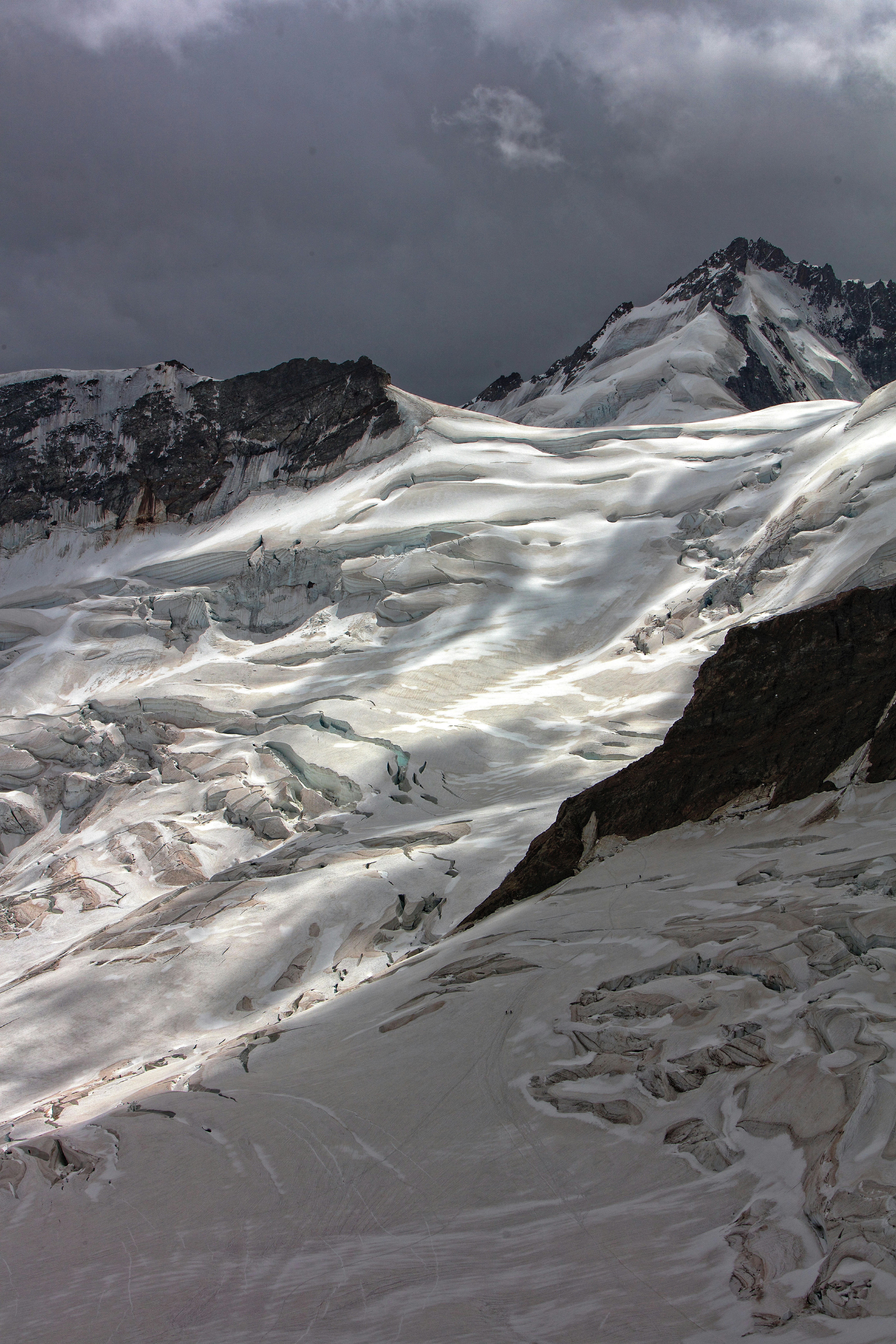 Intricate patterns of snow and ice weave through a majestic glacier under a moody sky. The scene captures the raw beauty of untouched alpine landscapes.