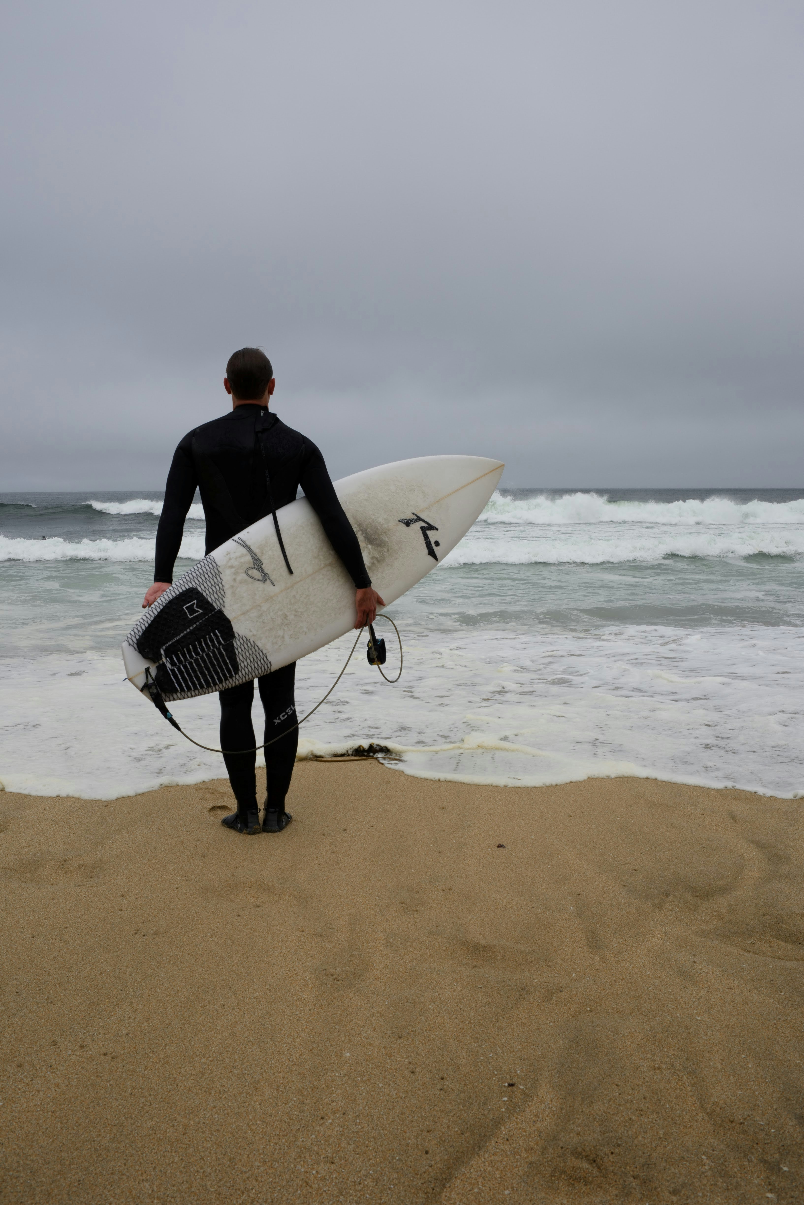 Surfer in a wetsuit gazes out at the ocean waves while holding a surfboard, set against a cloudy sky.
