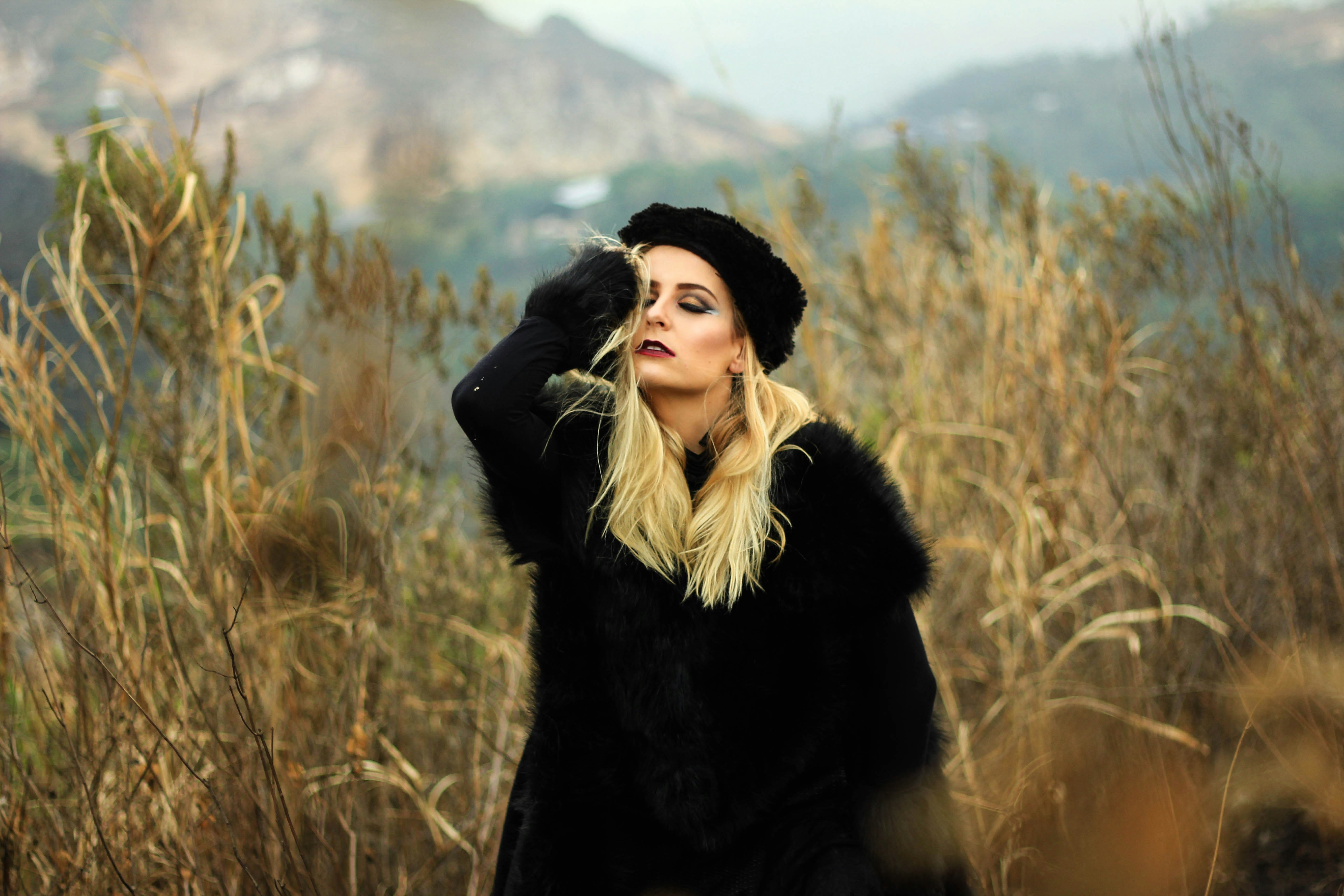 A fashionable woman in a black fur coat and hat poses amidst tall grass in a serene landscape. Her contemplative expression enhances the autumnal ambiance.