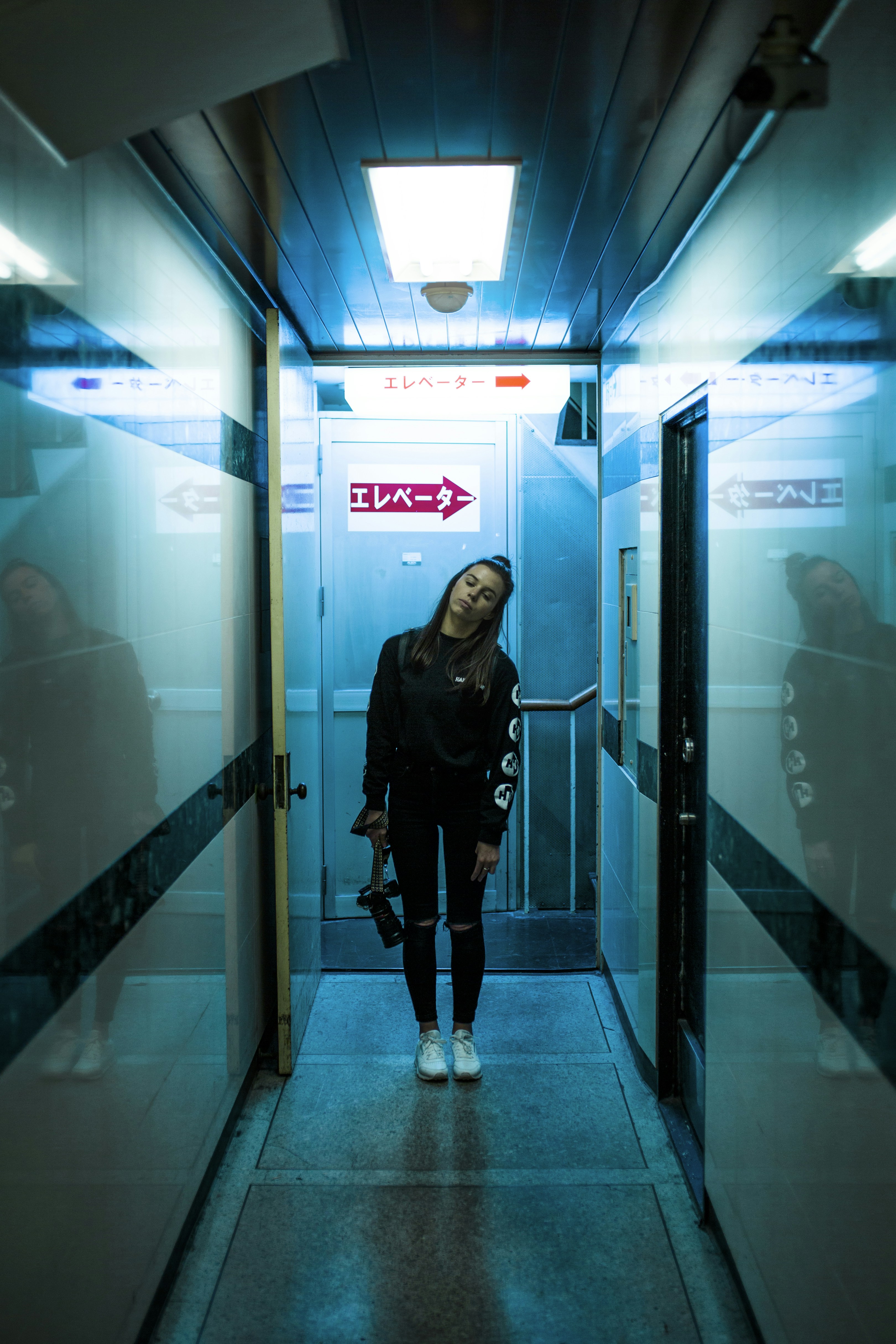 A young woman stands in a narrow corridor, holding a camera, with reflections on the glass walls and elevator signage above her. The atmosphere is tinged with a sense of quiet contemplation.