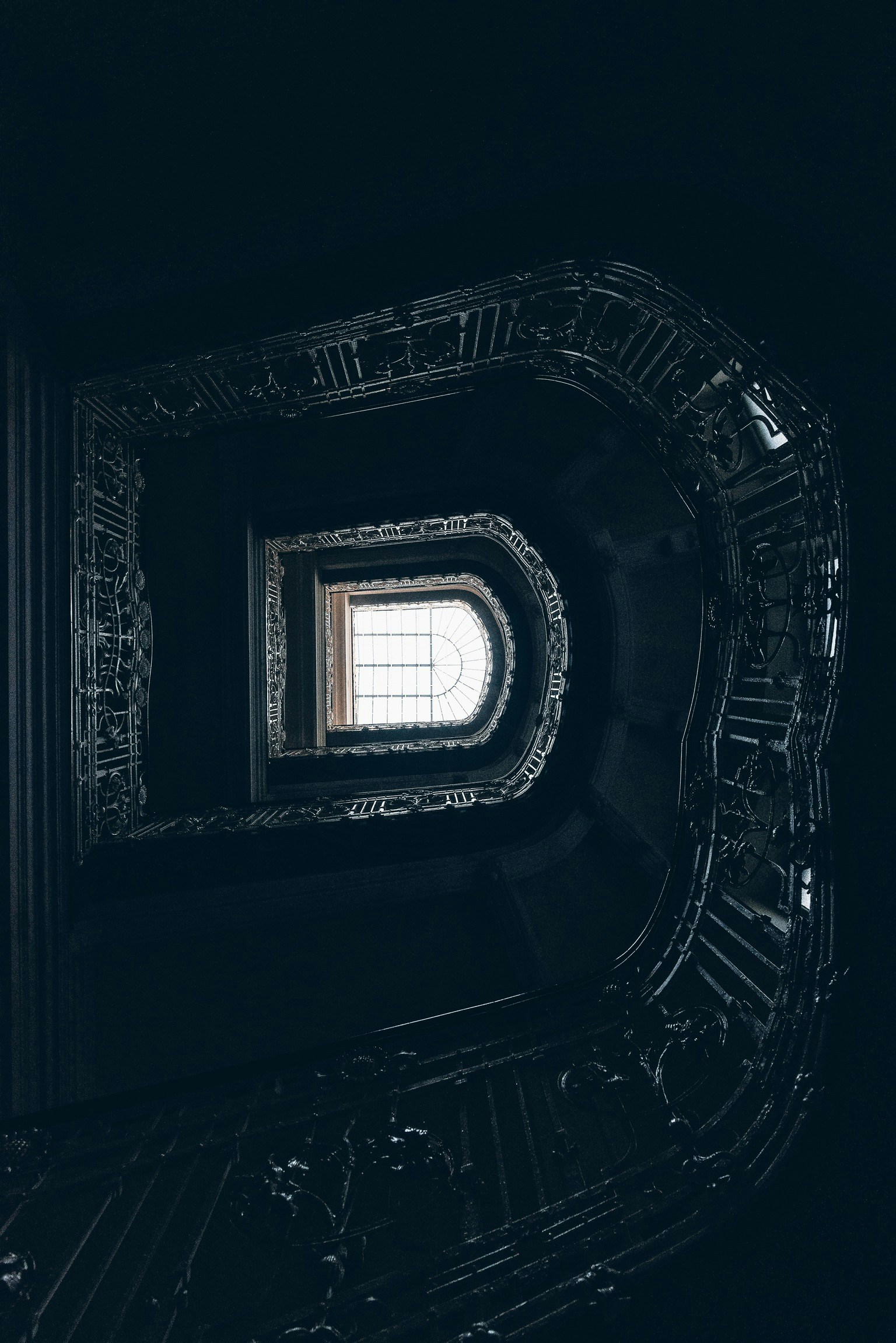 Ornate staircase spiraling upward, illuminated by a stained glass window at the top, creating a dramatic contrast with the surrounding darkness.