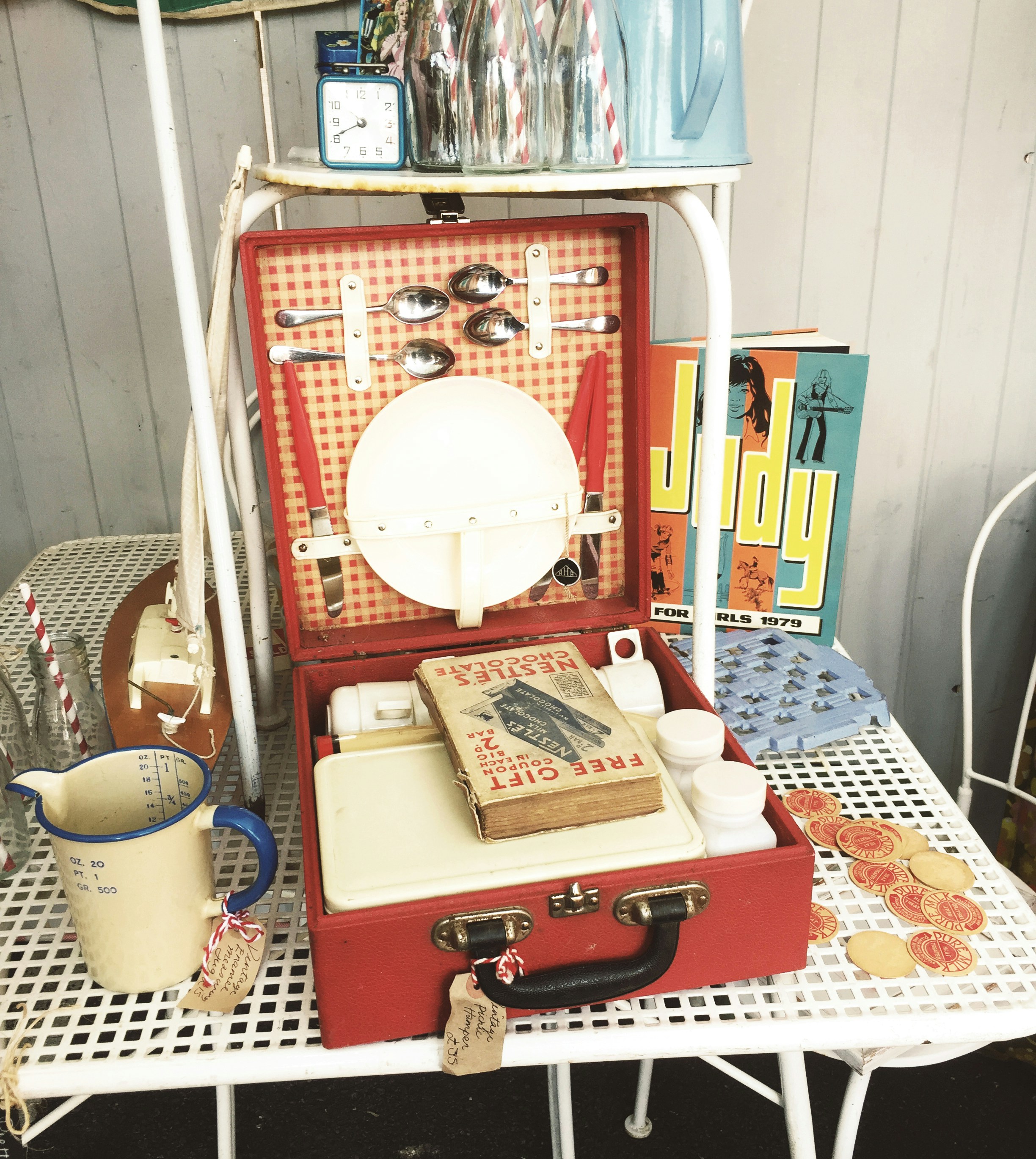 A retro suitcase filled with kitchen utensils, a measuring cup, and a vintage book, surrounded by charming collectibles on a white table.