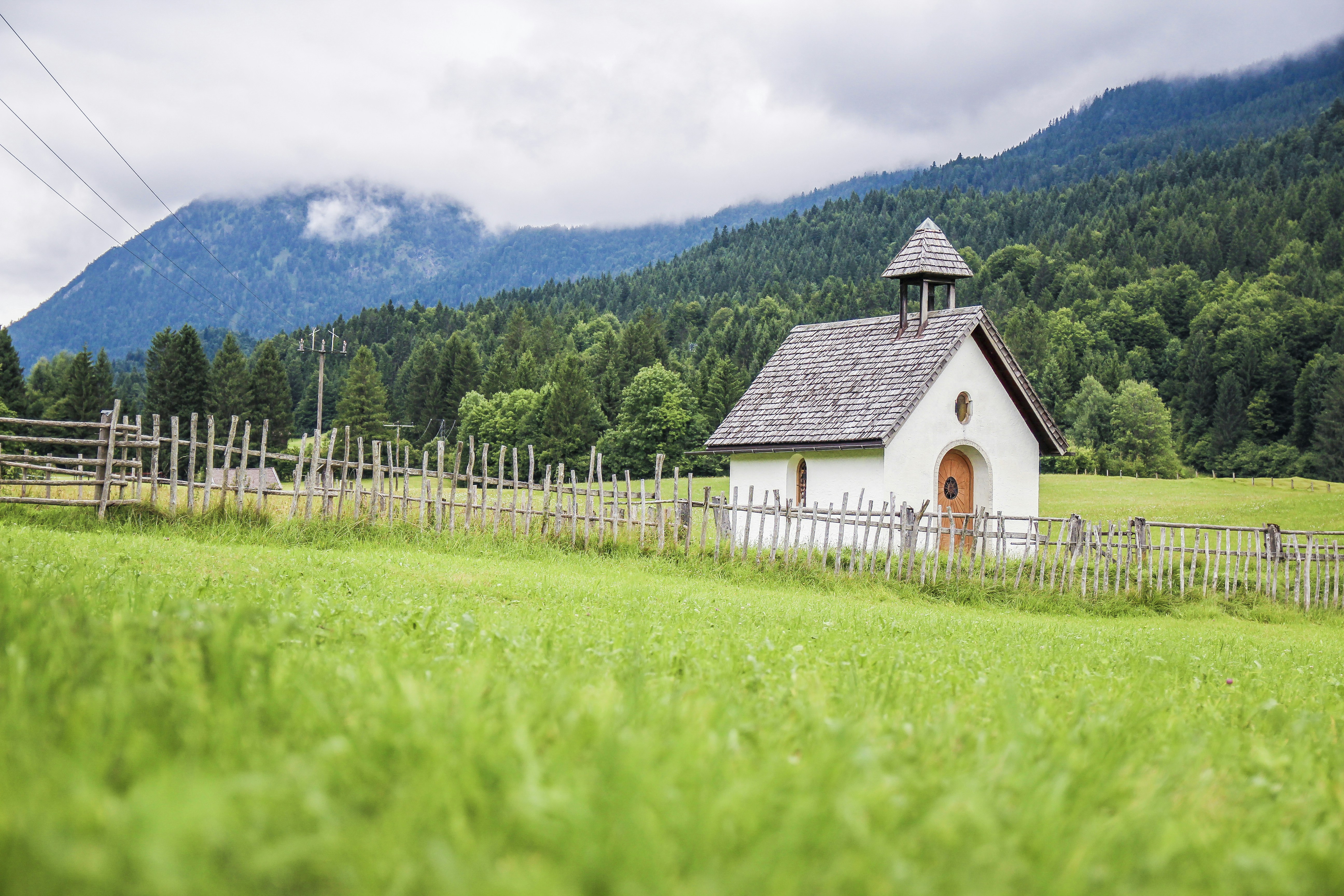 White chapel with a gray roof nestled in a green meadow, surrounded by a wooden fence and distant mountains.