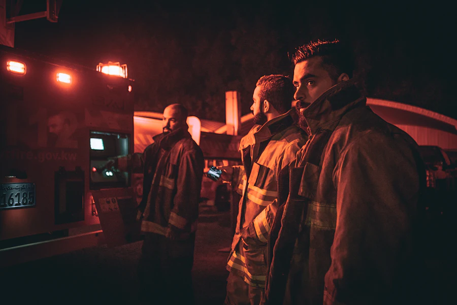 Three firefighters stand beside an emergency vehicle at night, lit by intense red lights. A fourth figure and the truck’s side details fade into the dark background.