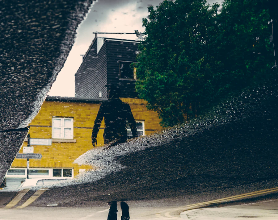Reflection of a person walking on a wet street beside a yellow building and greenery.