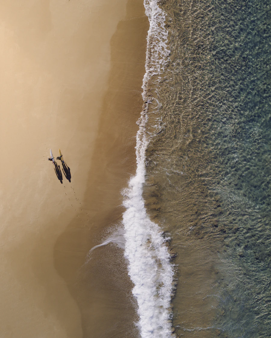 Aerial view of two surfers walking across a wide sandy beach beside foamy waves and turquoise water. Their long shadows stretch across the warm sand.