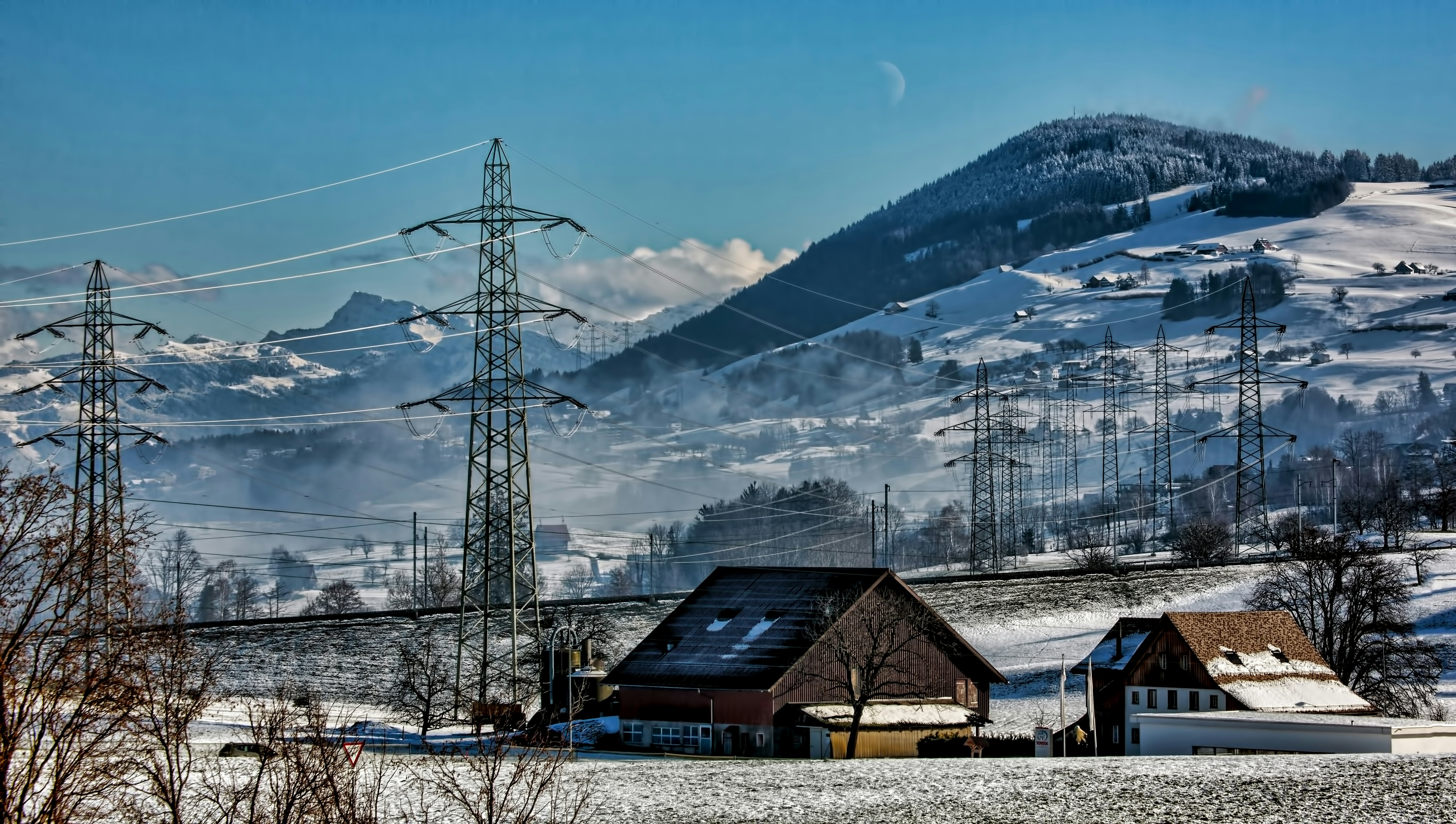 Snow-covered hills with power lines stretching across the frame, featuring rustic farmhouses in the foreground. The moon is faintly visible in the clear blue sky.