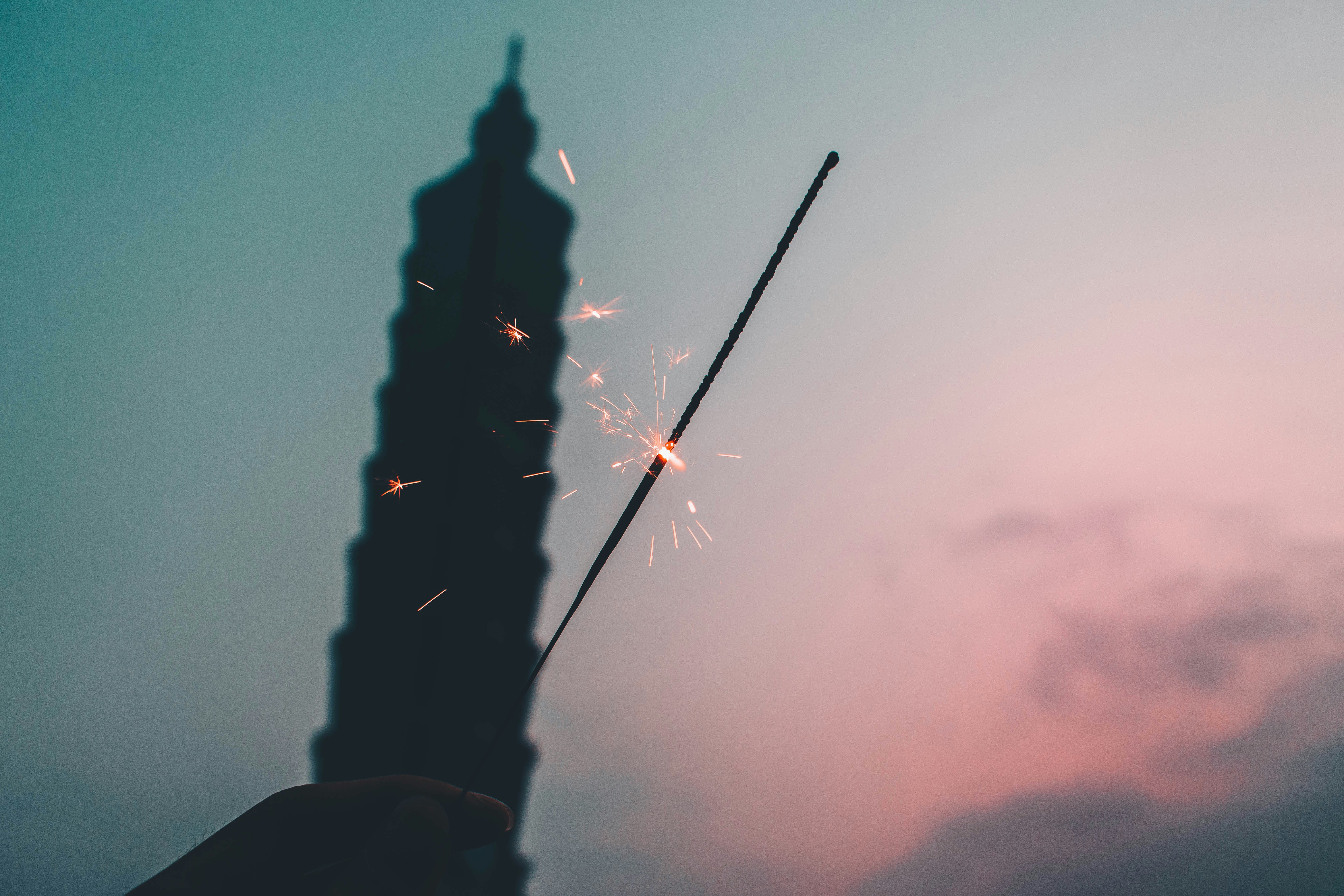 A hand holds a sparkler, illuminating the dusk sky while a tower looms in the background, creating a striking contrast between light and shadow.