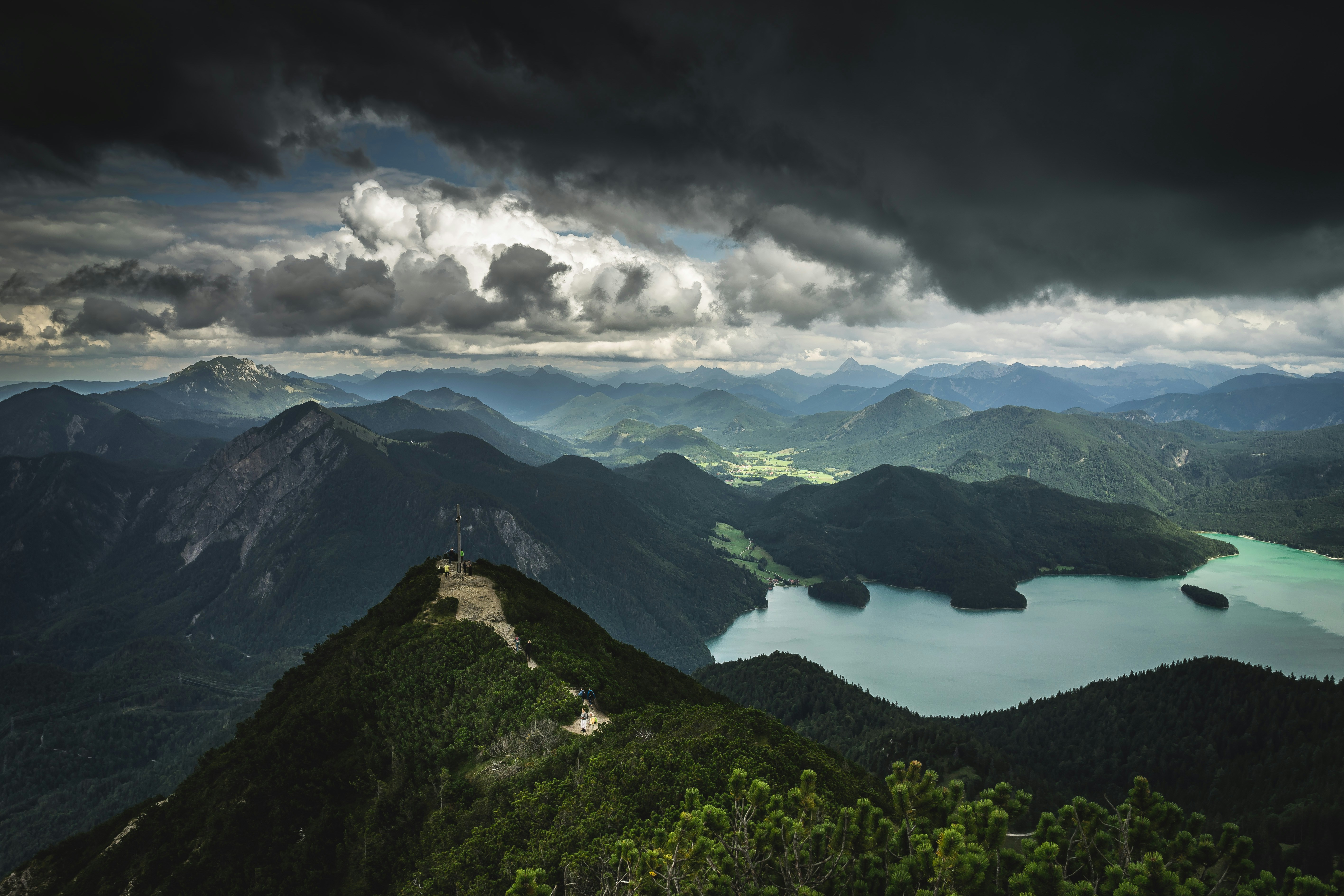 Dramatic clouds hover over a mountain ridge and serene lake surrounded by lush greenery.