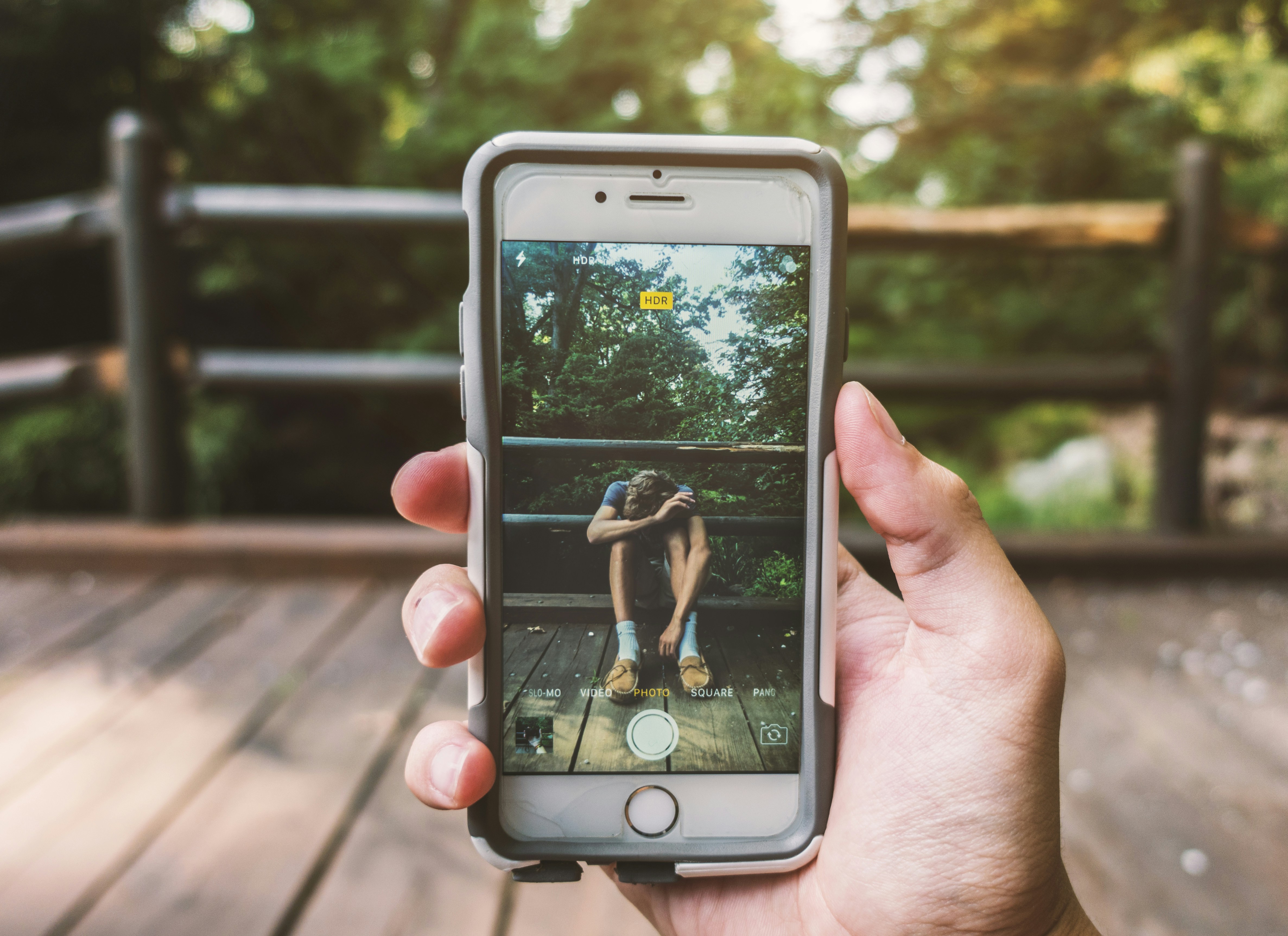 A hand holding a smartphone displaying a person sitting pensively on a wooden deck surrounded by greenery. The scene captures a moment of introspection.